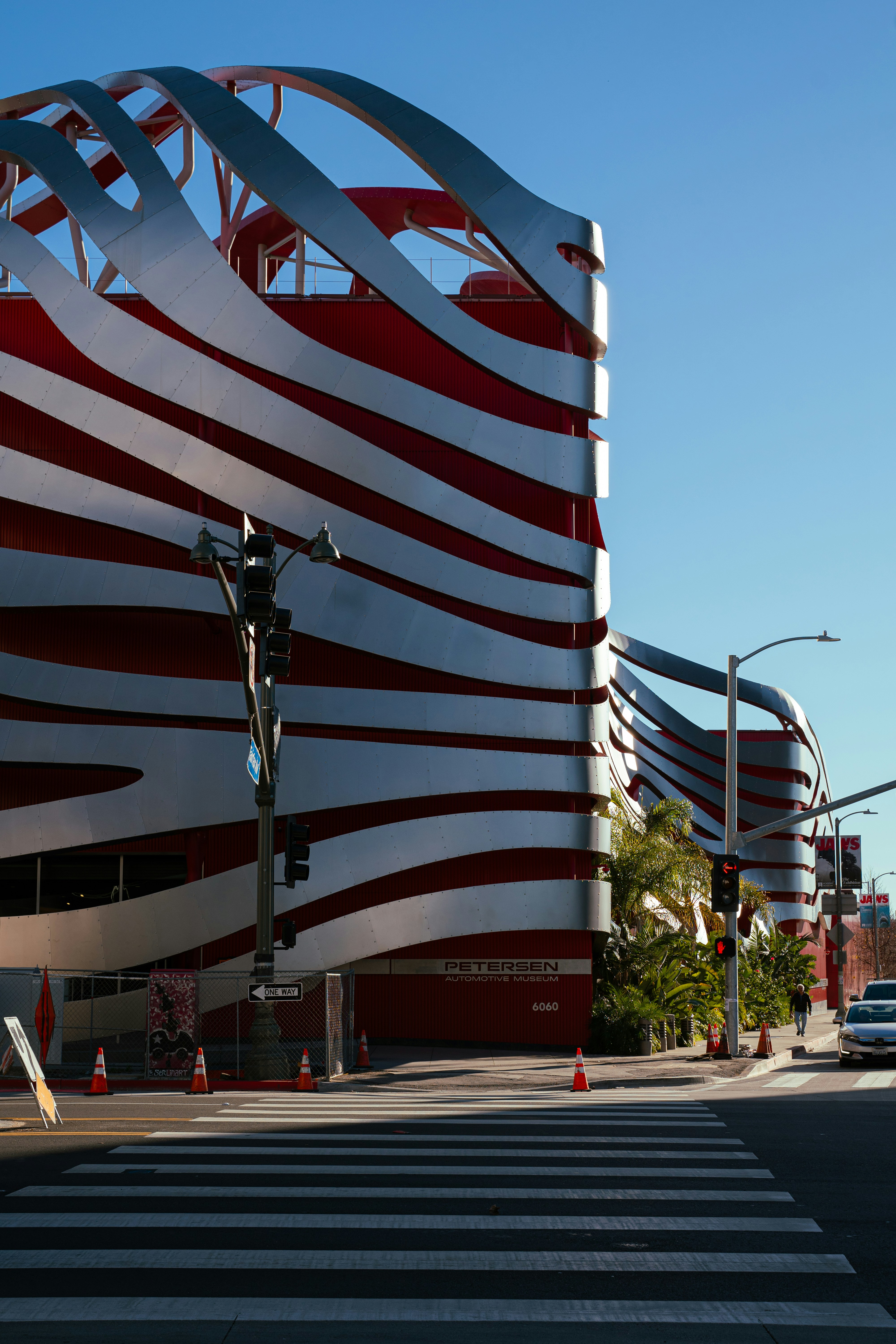 Architectural wave-pattern building along a sunlit street with crosswalk and traffic cones.