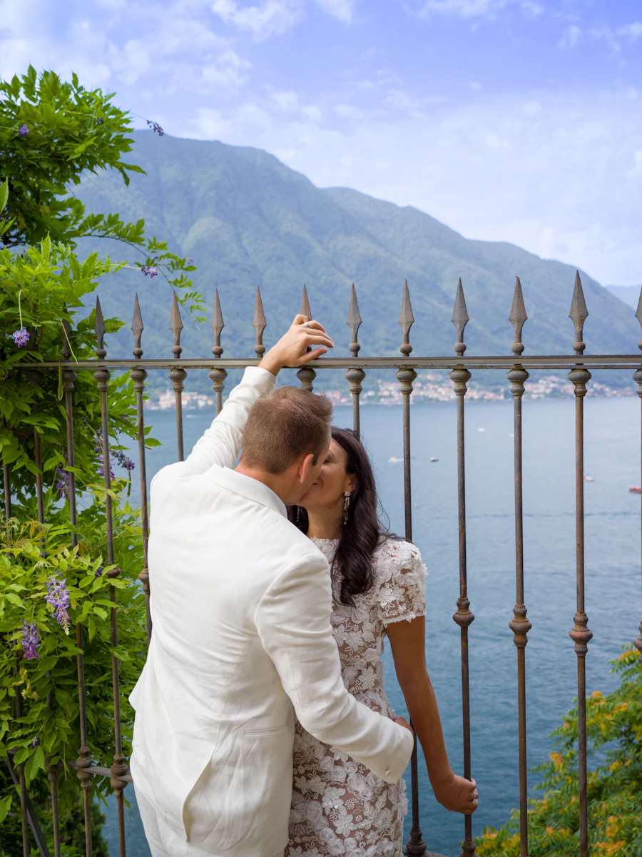 Couple sharing a kiss by a wrought-iron railing overlooking a lake with mountains in the distance; a garden frame surrounds them, suggesting a romantic outdoor setting. The scene is clearly a candid moment during a wedding or engagement shoot.