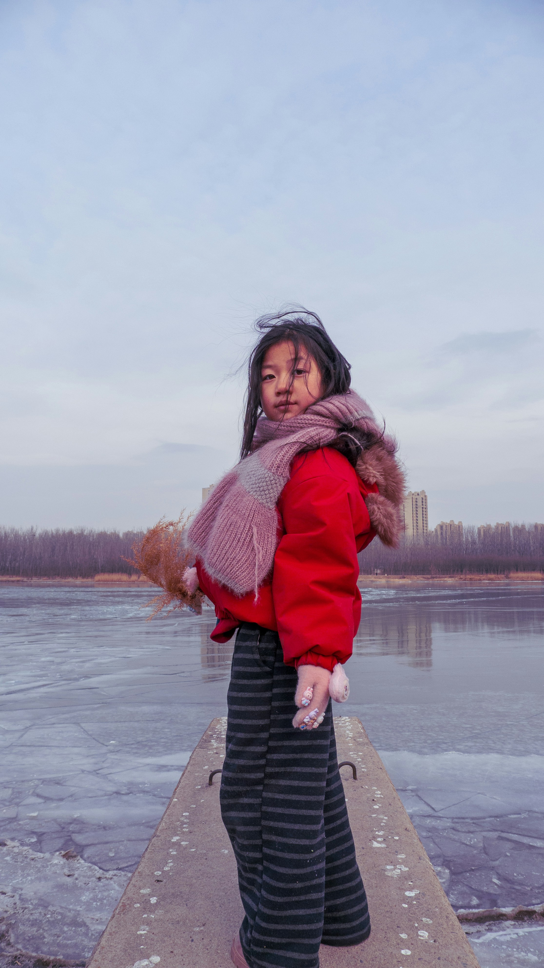 Young girl in a vibrant red jacket and fluffy pink scarf stands on a pier by a frozen lake, holding a dried plant—overcast winter sky, bare trees, and distant city buildings in the soft, serene background.
