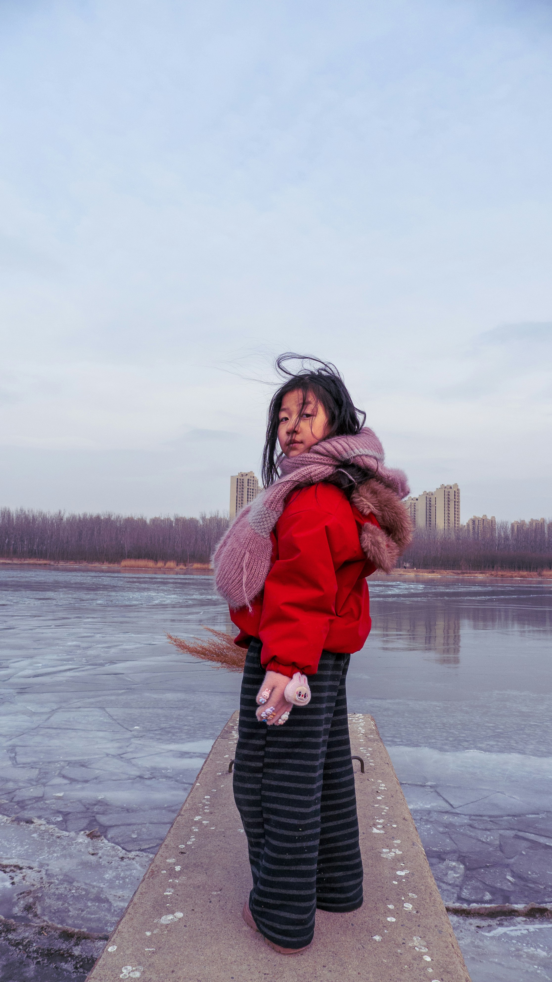 Young girl in a bright red jacket and pink scarf stands on a pier by a frozen lake, with windblown hair, city buildings and bare trees in the background under a soft overcast sky.