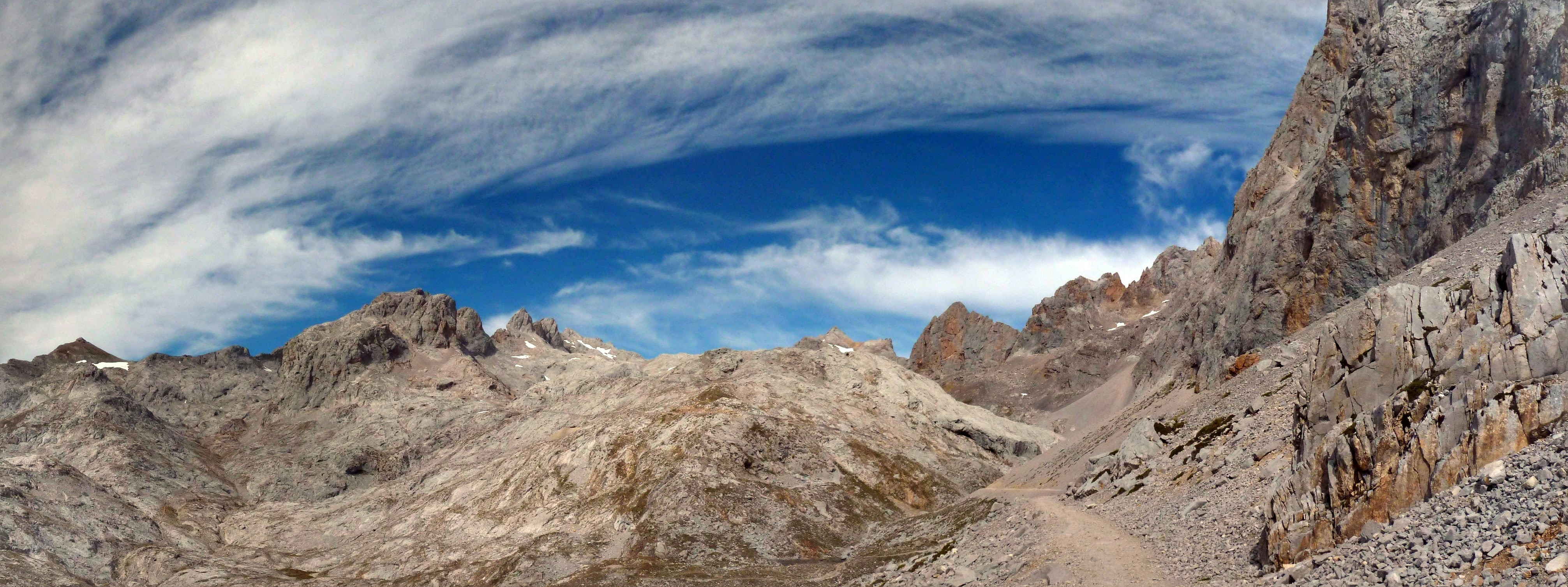 Majestic jagged mountains under a vivid blue sky with wispy clouds, showcasing stark rock textures and a wide-open alpine valley.