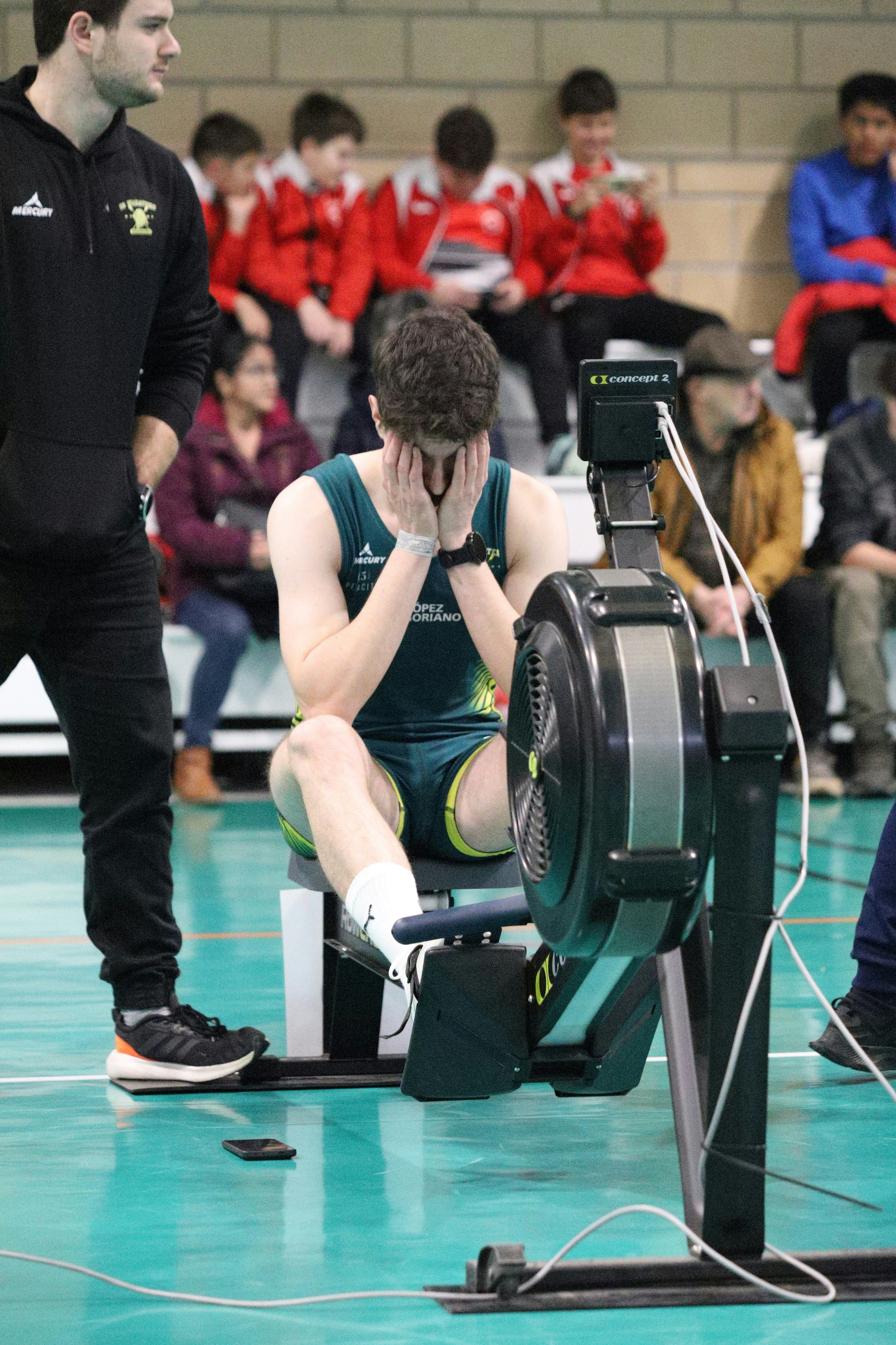Athlete in teal singlet covers his face while seated at a rowing machine in a gym, with a coach nearby and spectators in the background.