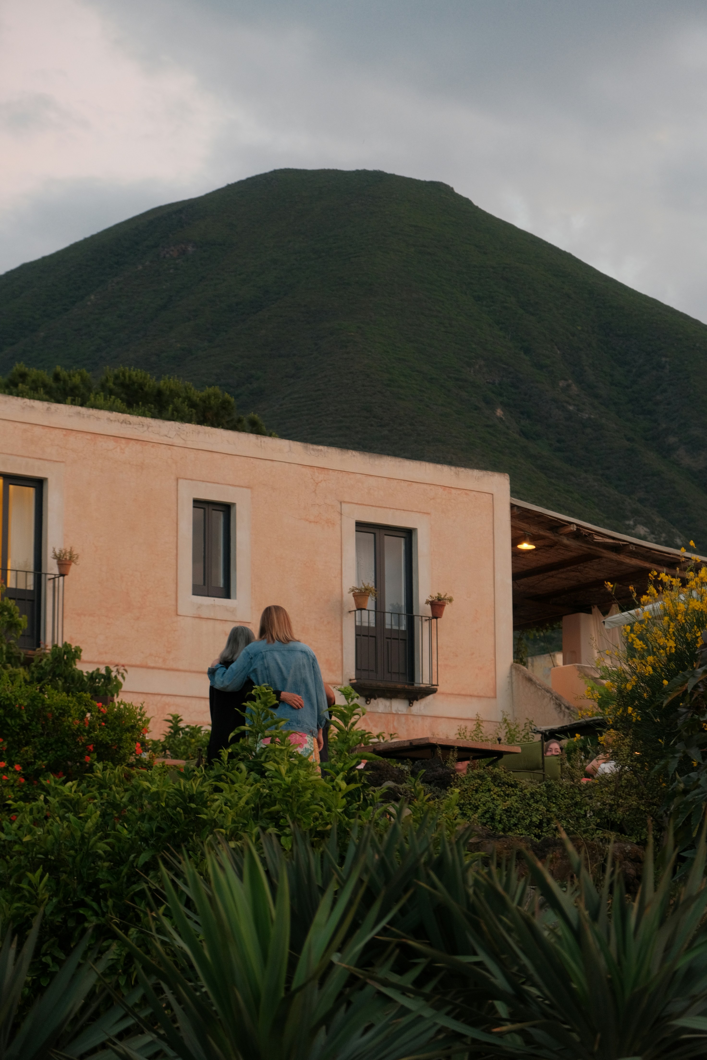 Two figures embrace at twilight beside a sunlit villa with a looming green hill