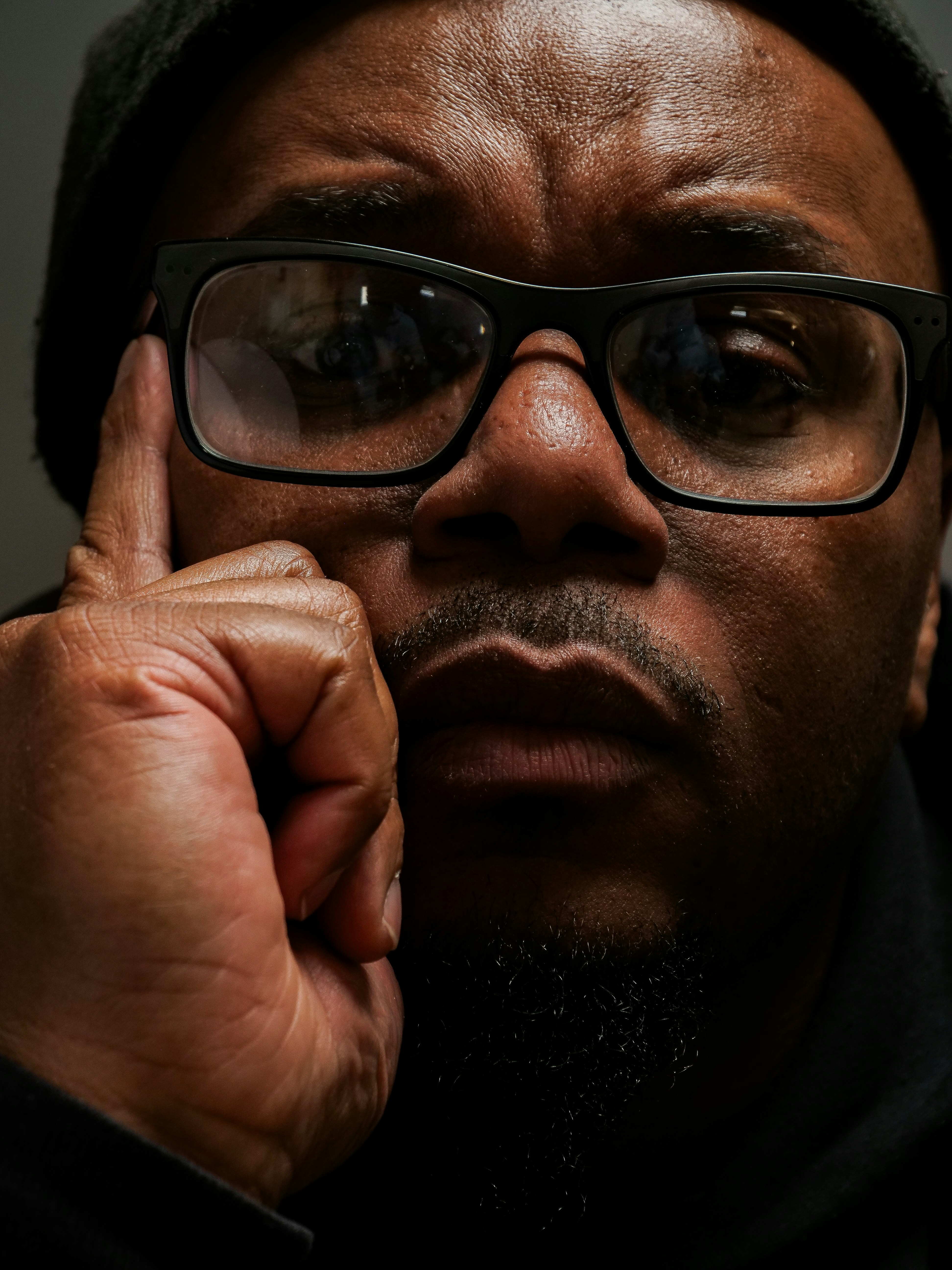 Close-up portrait with a man wearing glasses, resting his cheek on his finger; intimate, moody lighting highlighting facial textures and stubble; studio or indoor setting with subdued background.
