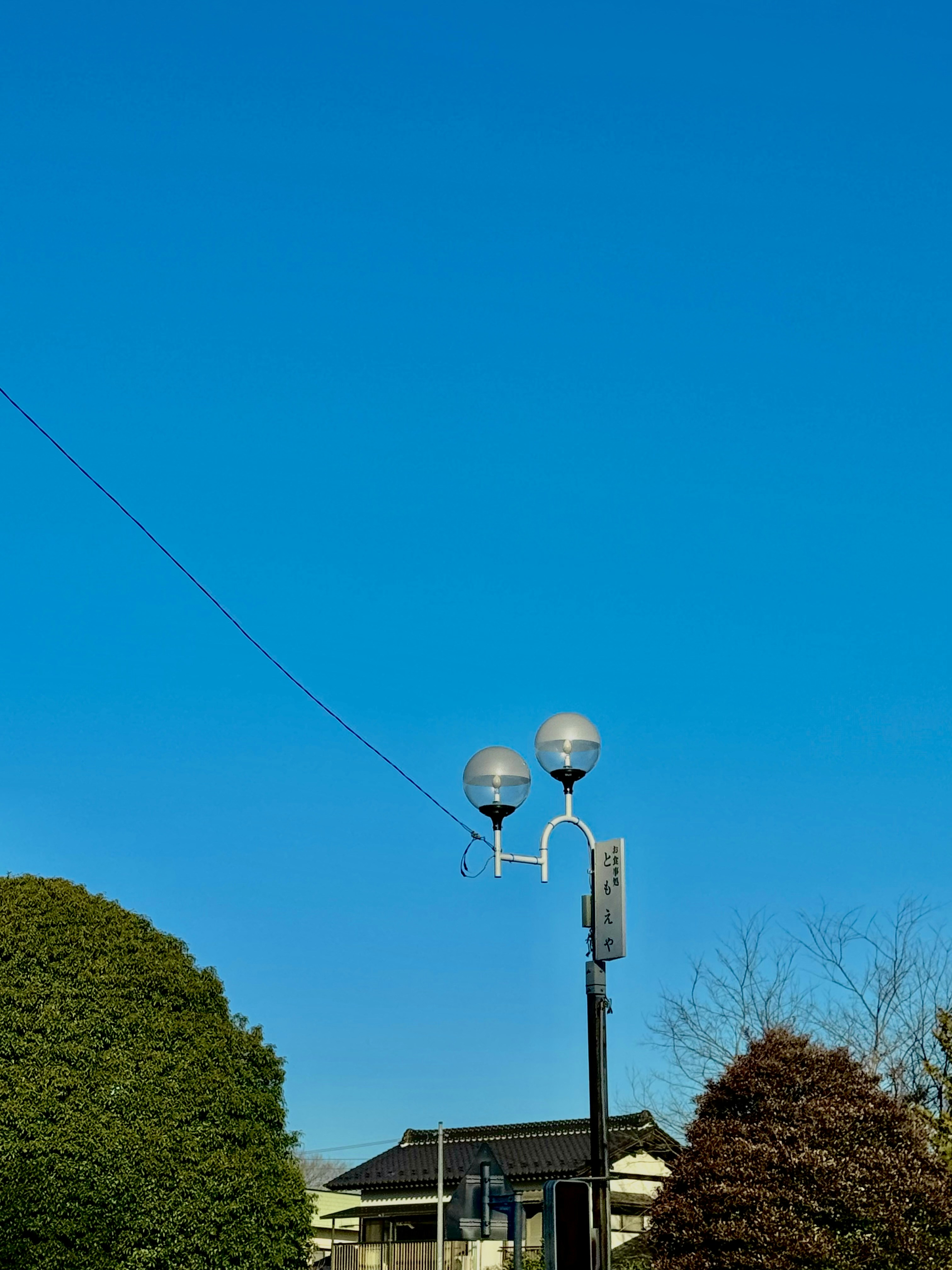Tall street lamp with two globes against a bright blue sky; lower scene shows trimmed greenery and a traditional building. A subtle power line arcs diagonally across the frame.