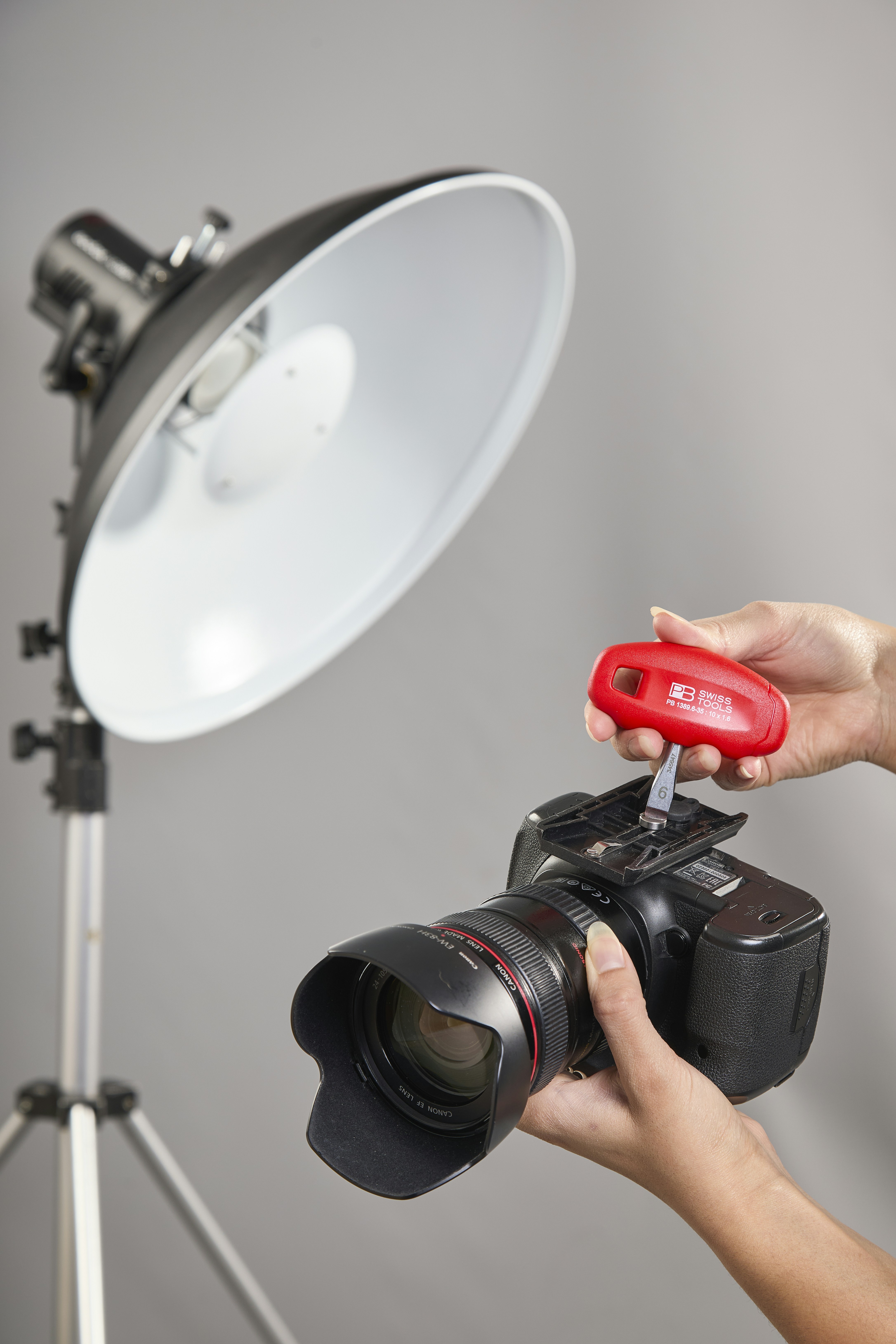 Close-up of a photographer's hands adjusting a red trigger atop a DSLR, with a large studio reflector on a stand in the background.