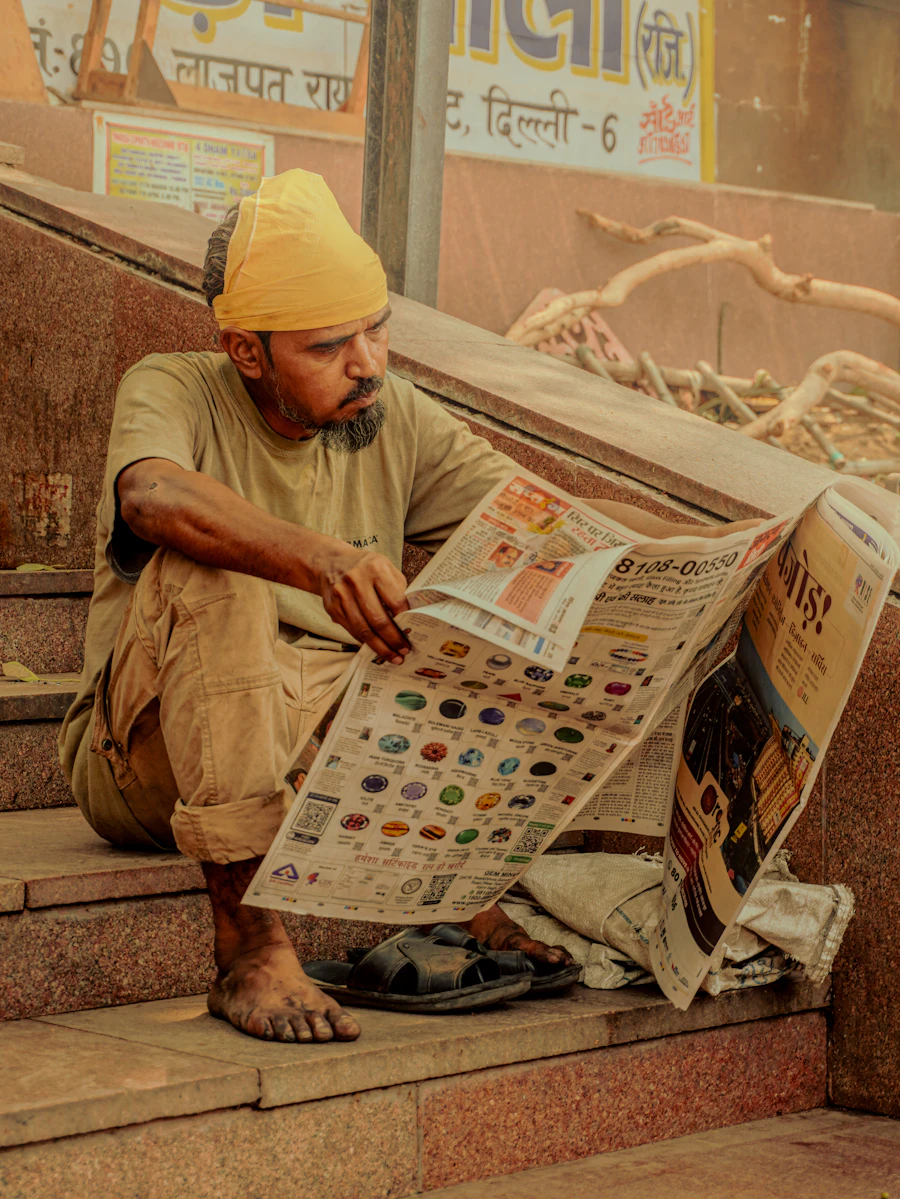 An Indian man in a yellow turban sits barefoot on stone steps, engrossed in a newspaper beside a parked sandal and a stack of papers, bathed in warm light.