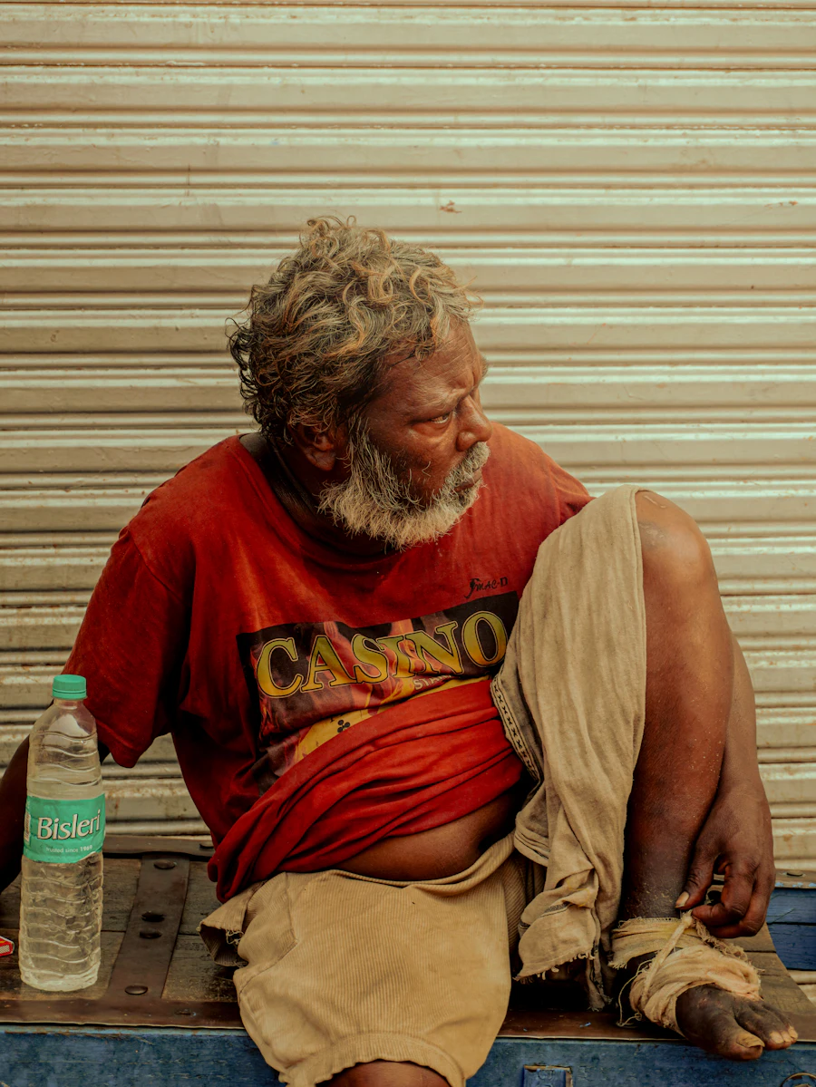 An older man with a red shirt sits on a platform beside a bottle of water, gazing off to the side; the scene captures everyday hardship and momentary respite in a sunlit urban setting. (Photograph) 