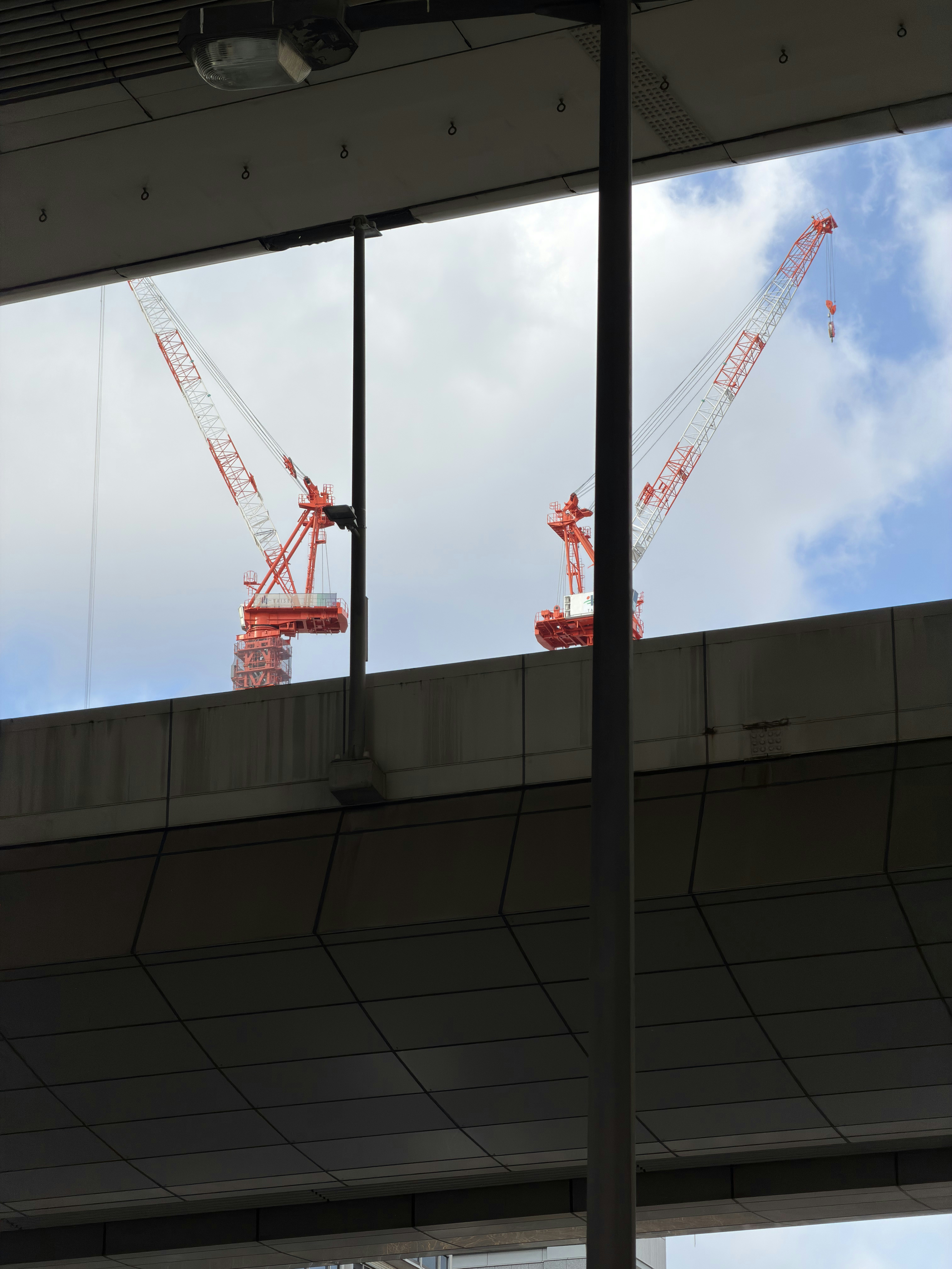 Red crane silhouettes pierce a blue-sky gap beneath a city-overpass