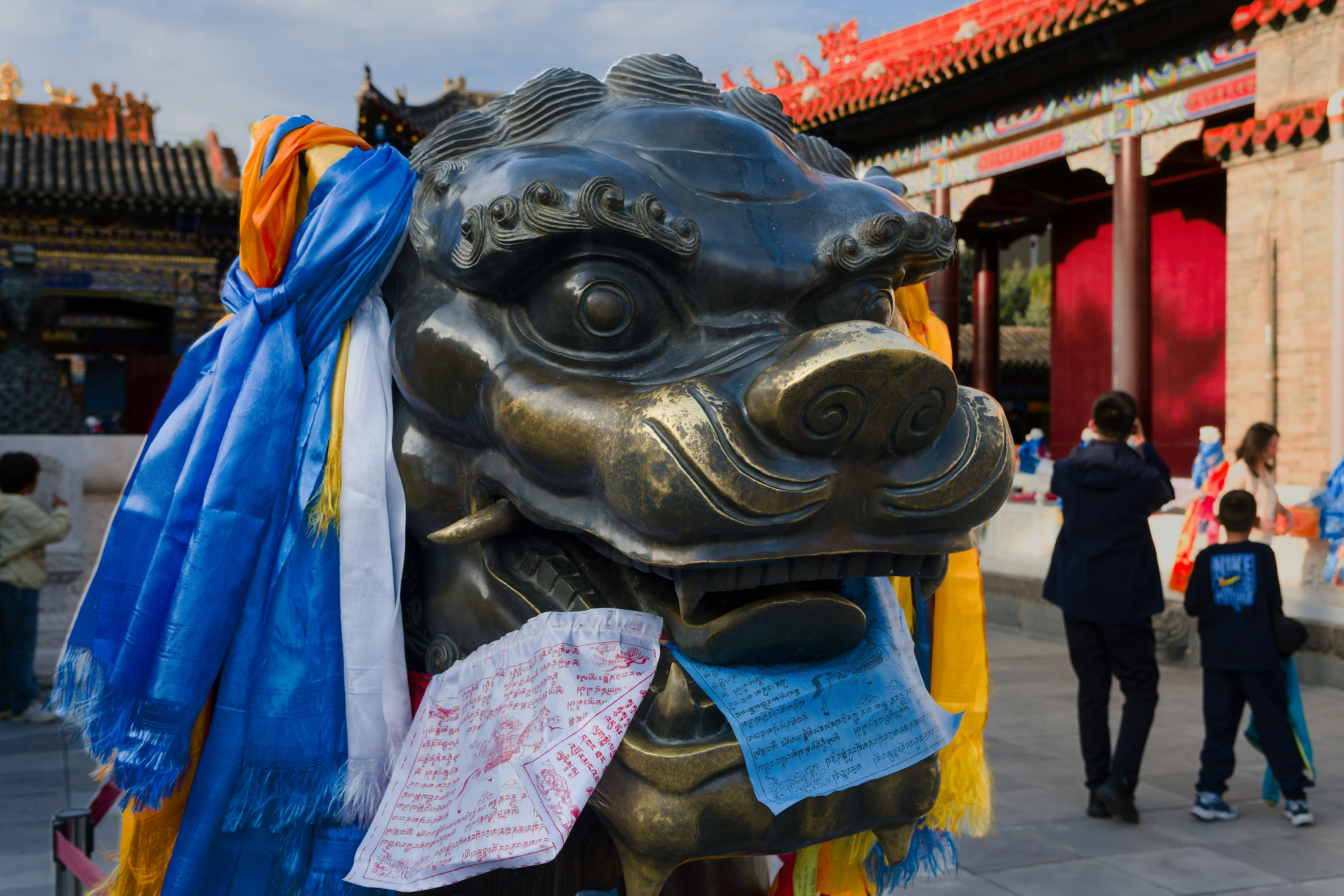 Metal lion sculpture with colorful cloth and prayer flags in an ancient temple setting during the day - A metal lion sculpture stands at the entrance of a temple. Colorful cloth and prayer flags hang from its mouth. People walk nearby, enjoying the sights. The temple has rich architecture.