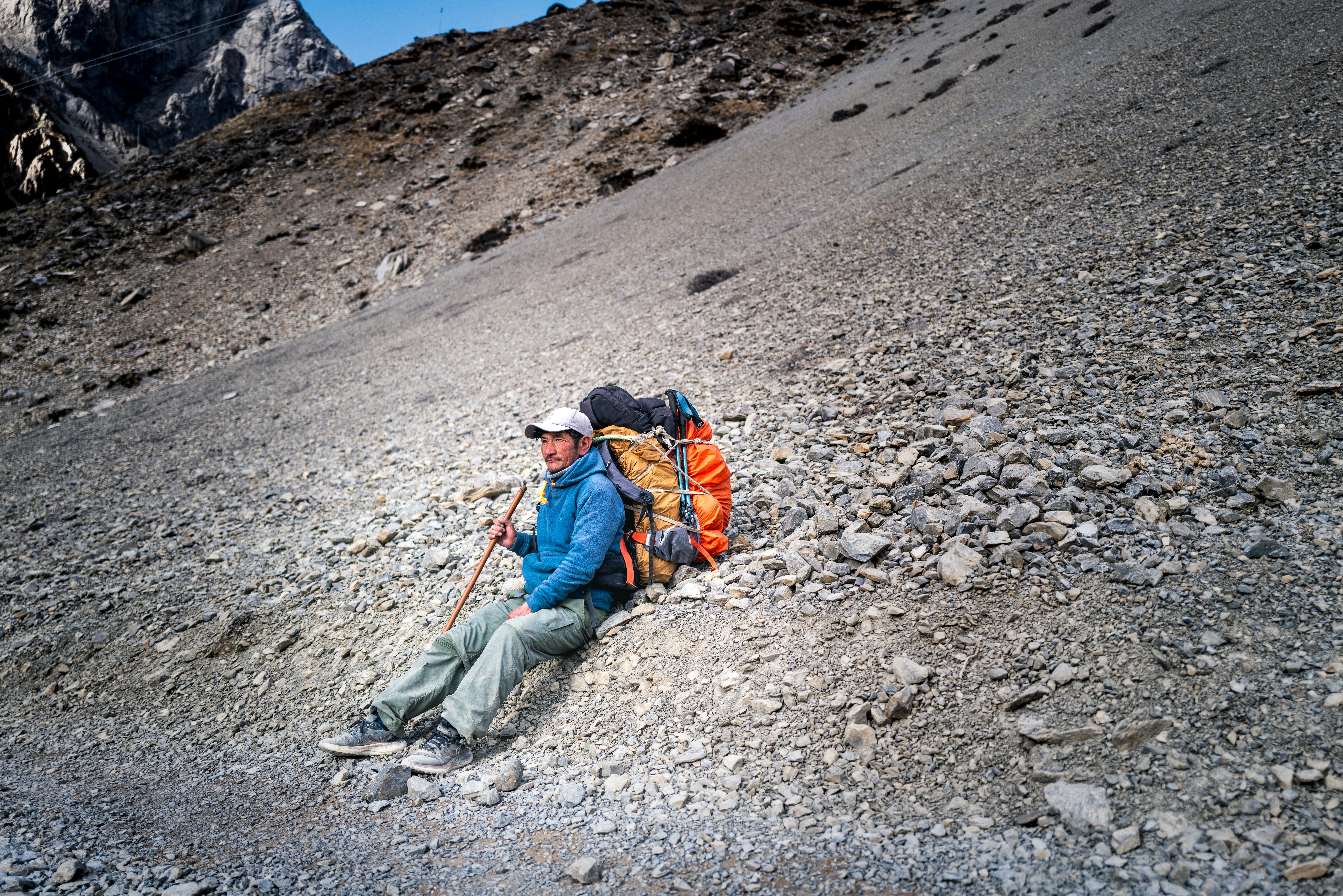 Trekking guide rests on a rocky Himalayan trail, equipped with a large orange backpack and wooden walking stick, against a rugged mountain slope under a bright blue sky — outdoor adventure, high-altitude hiking