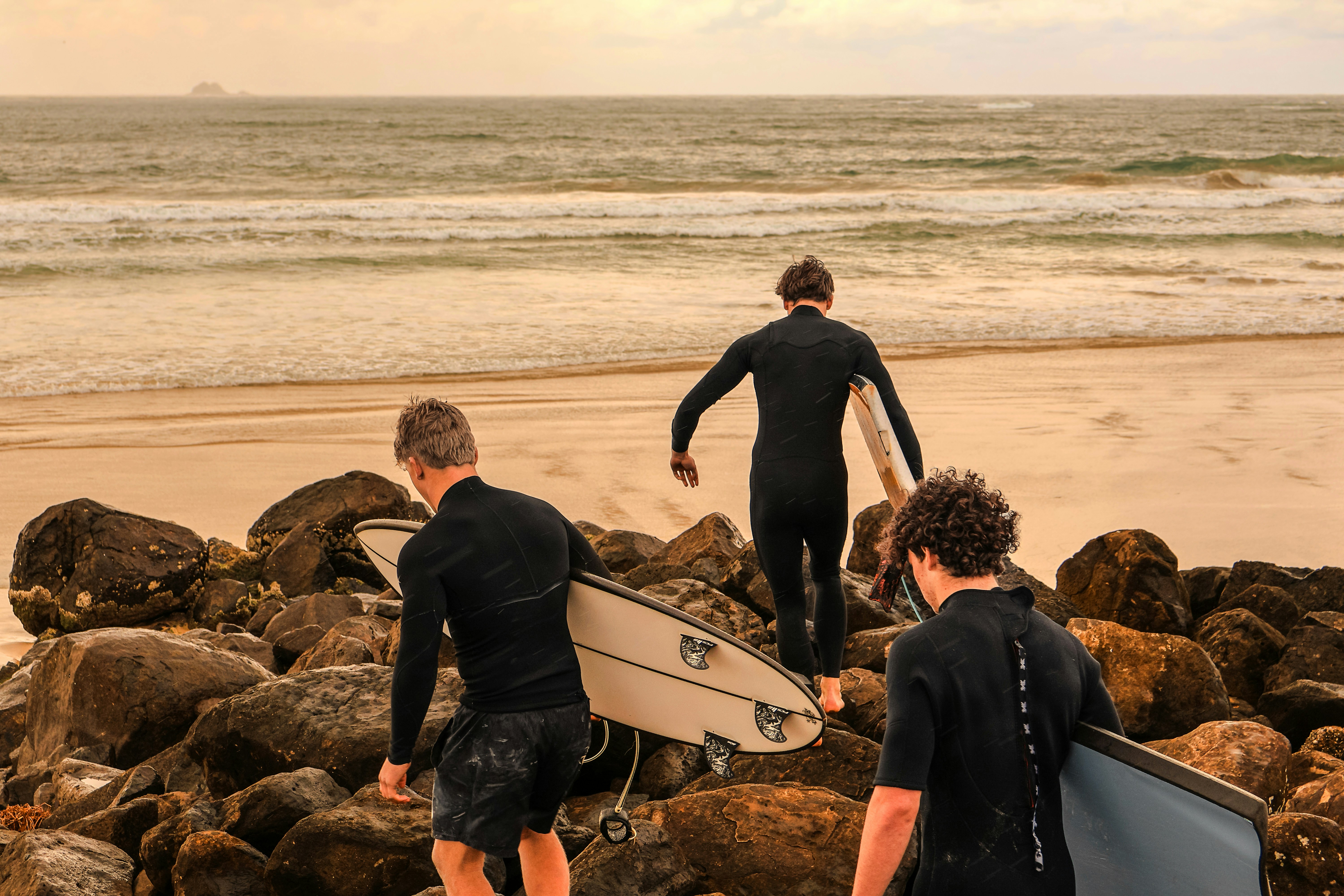Rising to the Shore: Trio of Surfers Converge on a rocky beach at dusk