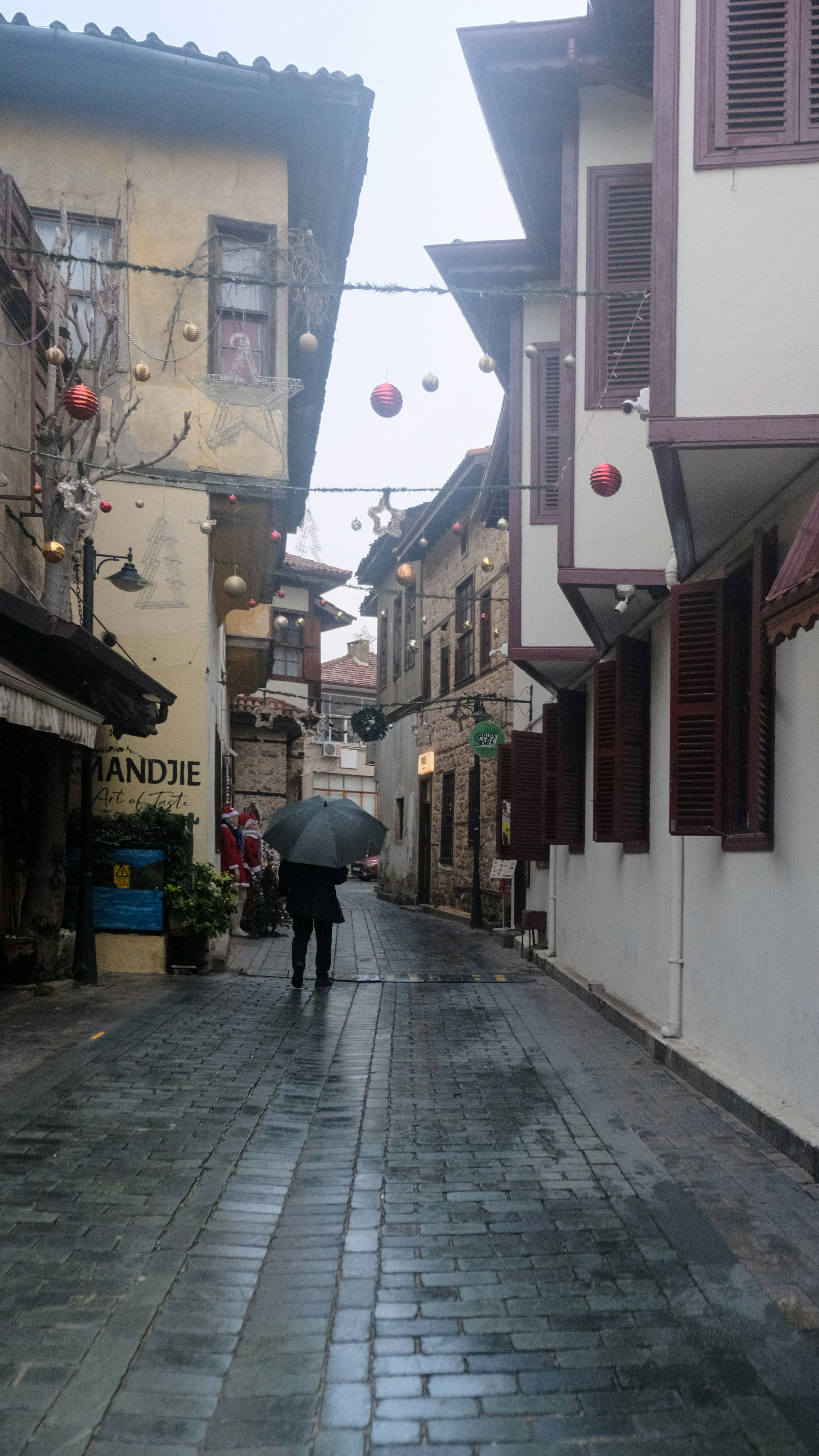 Cozy cobbled street under festive orbs, a lone figure with umbrella drifting toward the central plaza