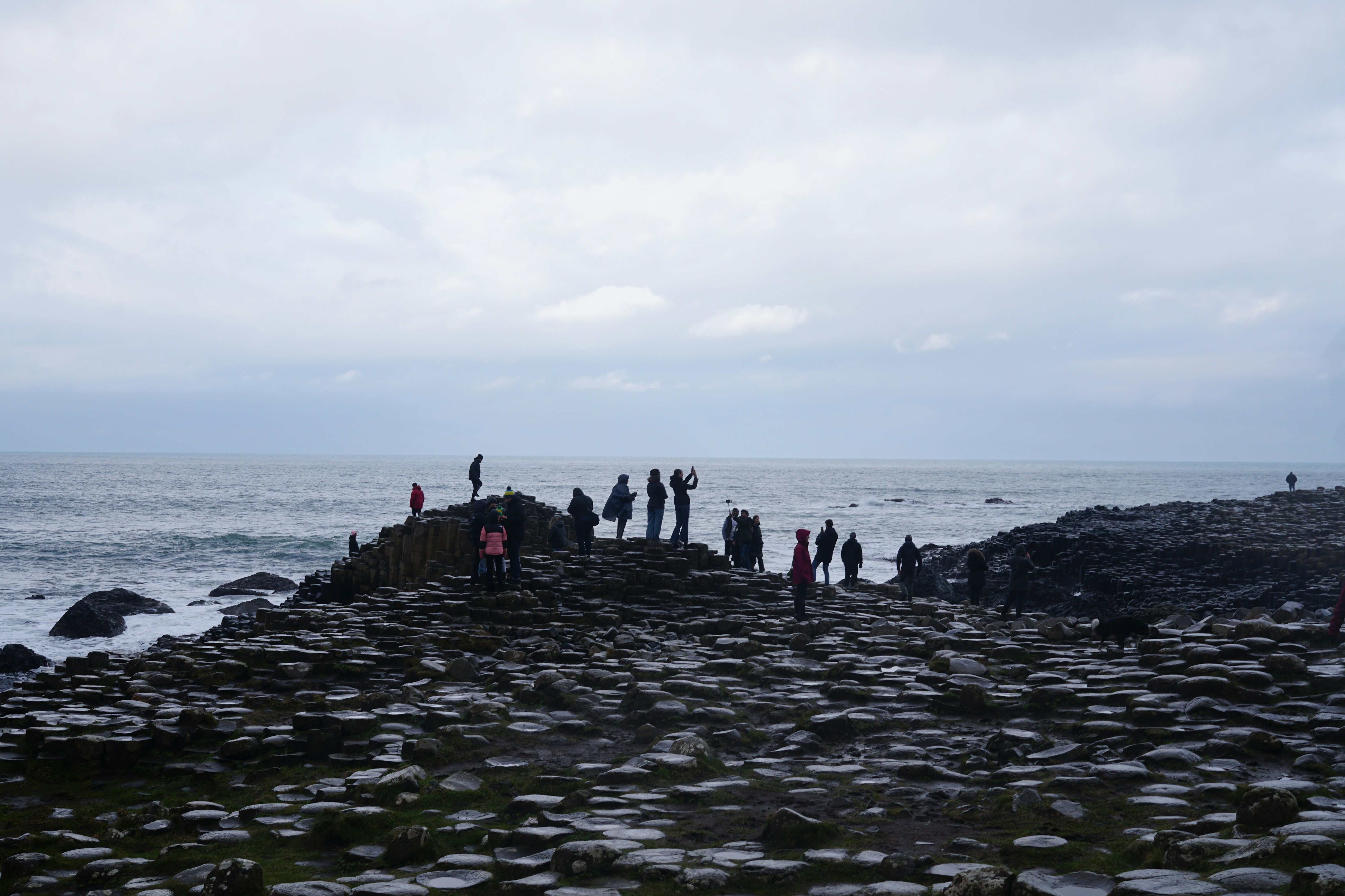 People on a rocky jetty extending into a calm sea under a soft cloudy sky