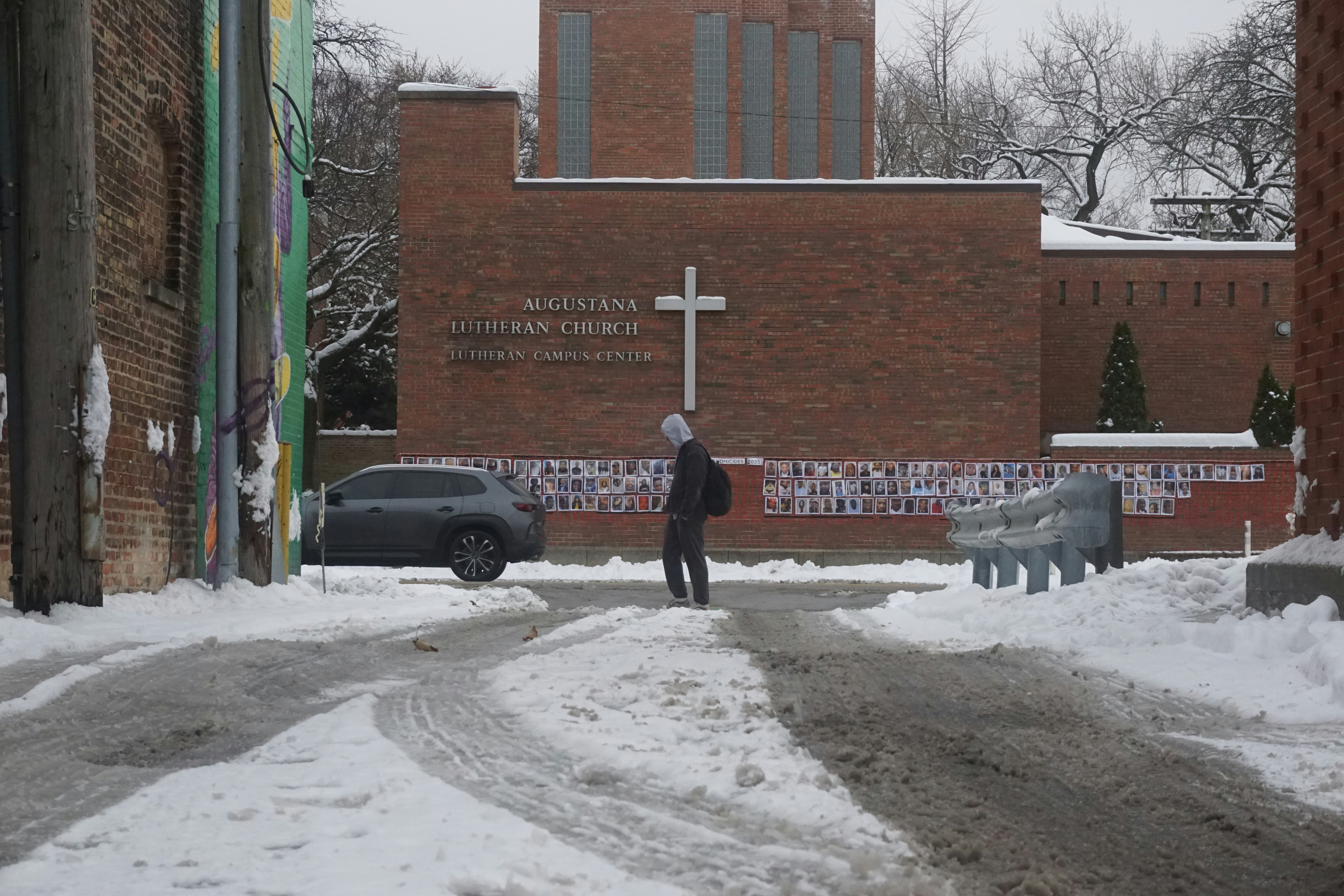 Snow-covered street view with a lone figure near a brick church, benches along the path and parked car, capturing quiet winter ambience.