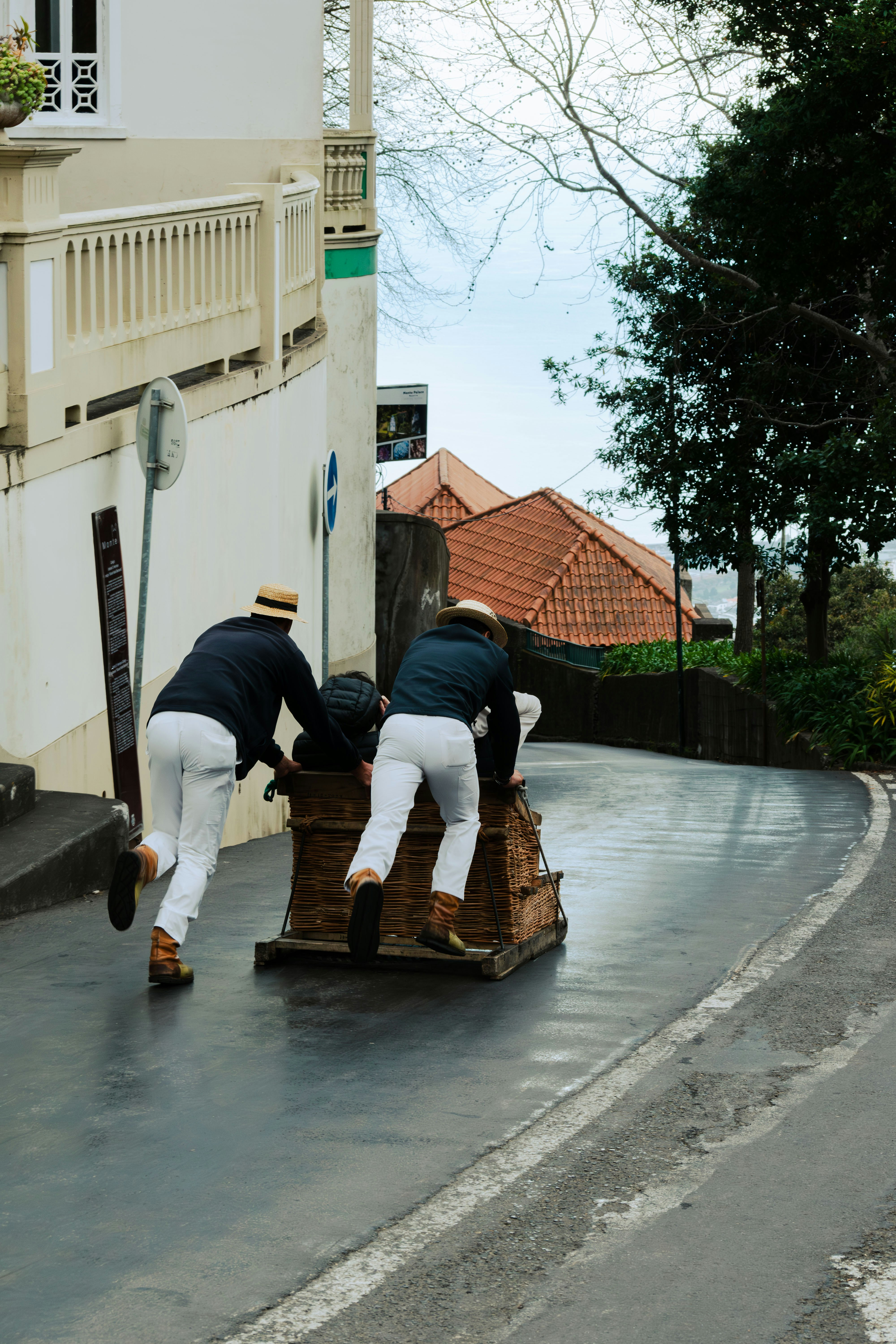 Three Movers on a Rainy Hill Carry a Woven Obstacle Through a Quiet Street