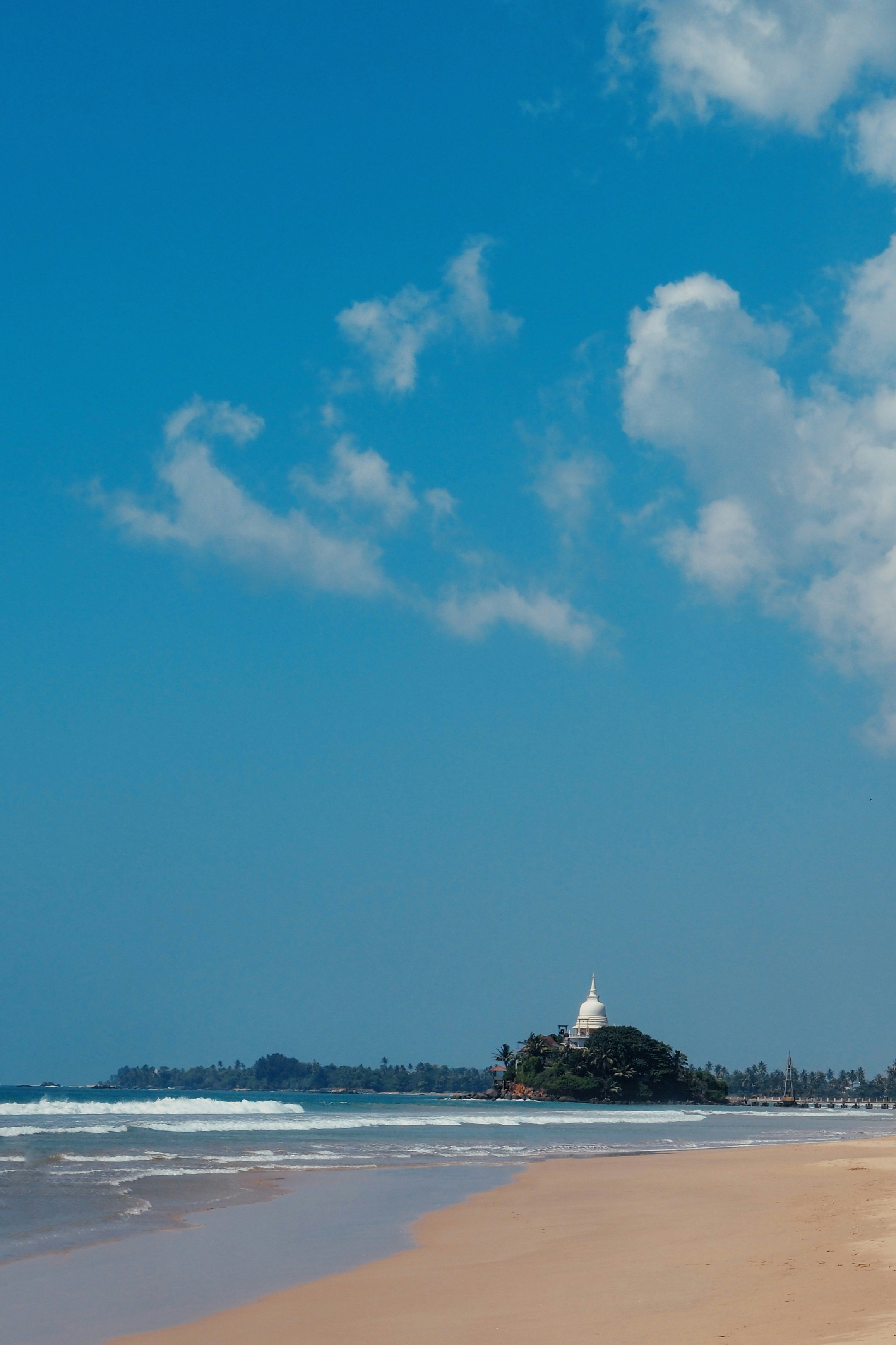 A serene beach scene with a distant white temple on a small island, waves lapping the sandy shore under a clear blue sky.