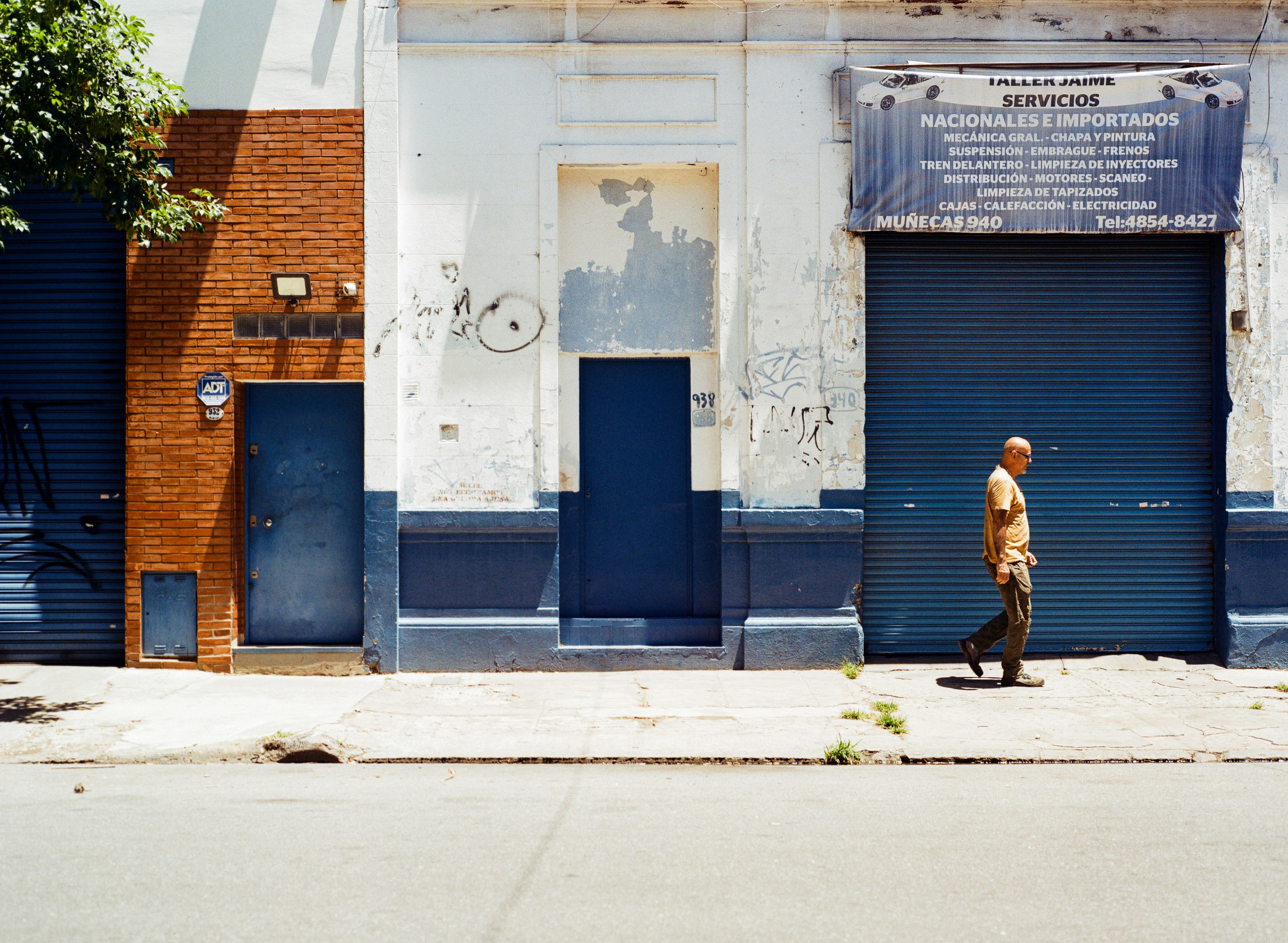 A man walking past closed storefronts on a quiet city street. Strong midday light and simple geometry create a minimalist street photography scene focused on rhythm, color, and urban life.