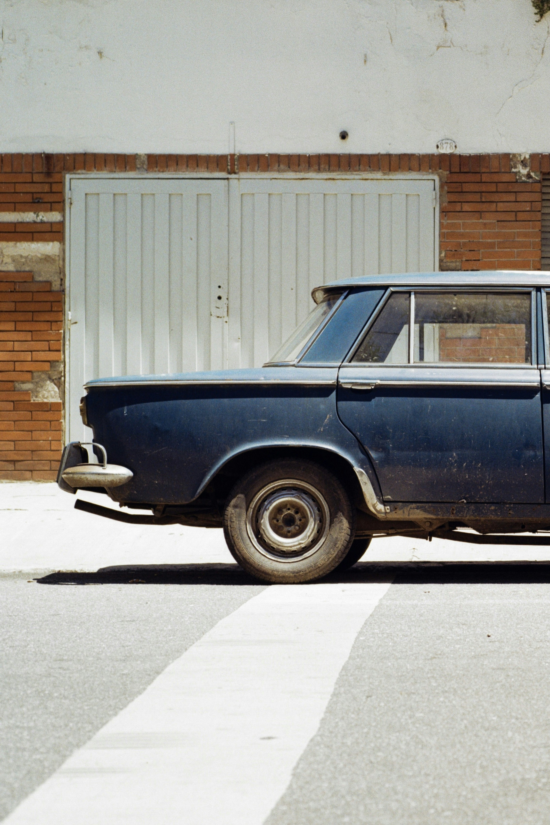A vintage blue car parked on a quiet street, framed by a pedestrian crossing and a sunlit wall. A simple urban scene capturing stillness, nostalgia, and everyday life in the city.