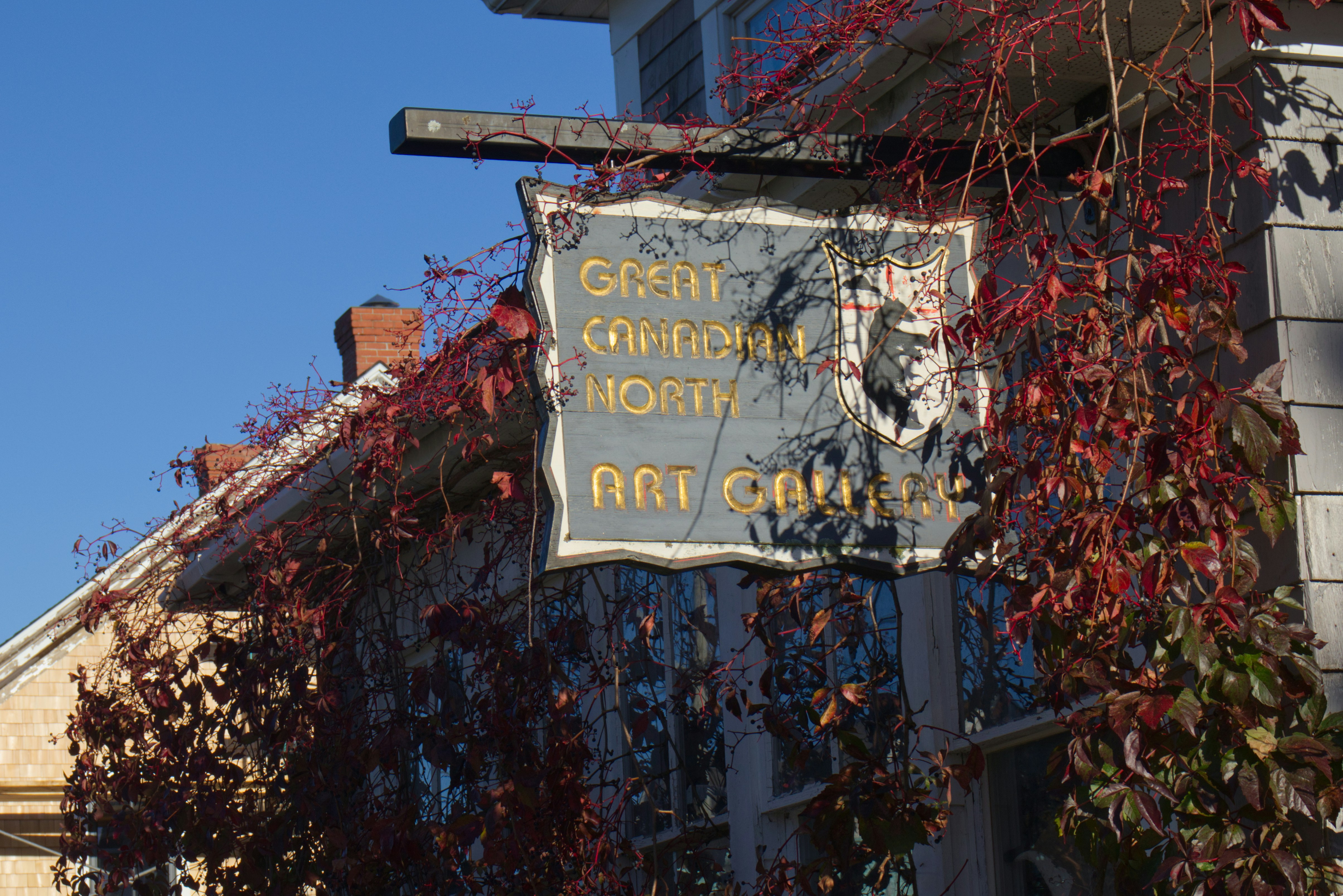Signboard of the Great Canadian North Art Gallery wrapped in autumn vines against a clear blue sky.