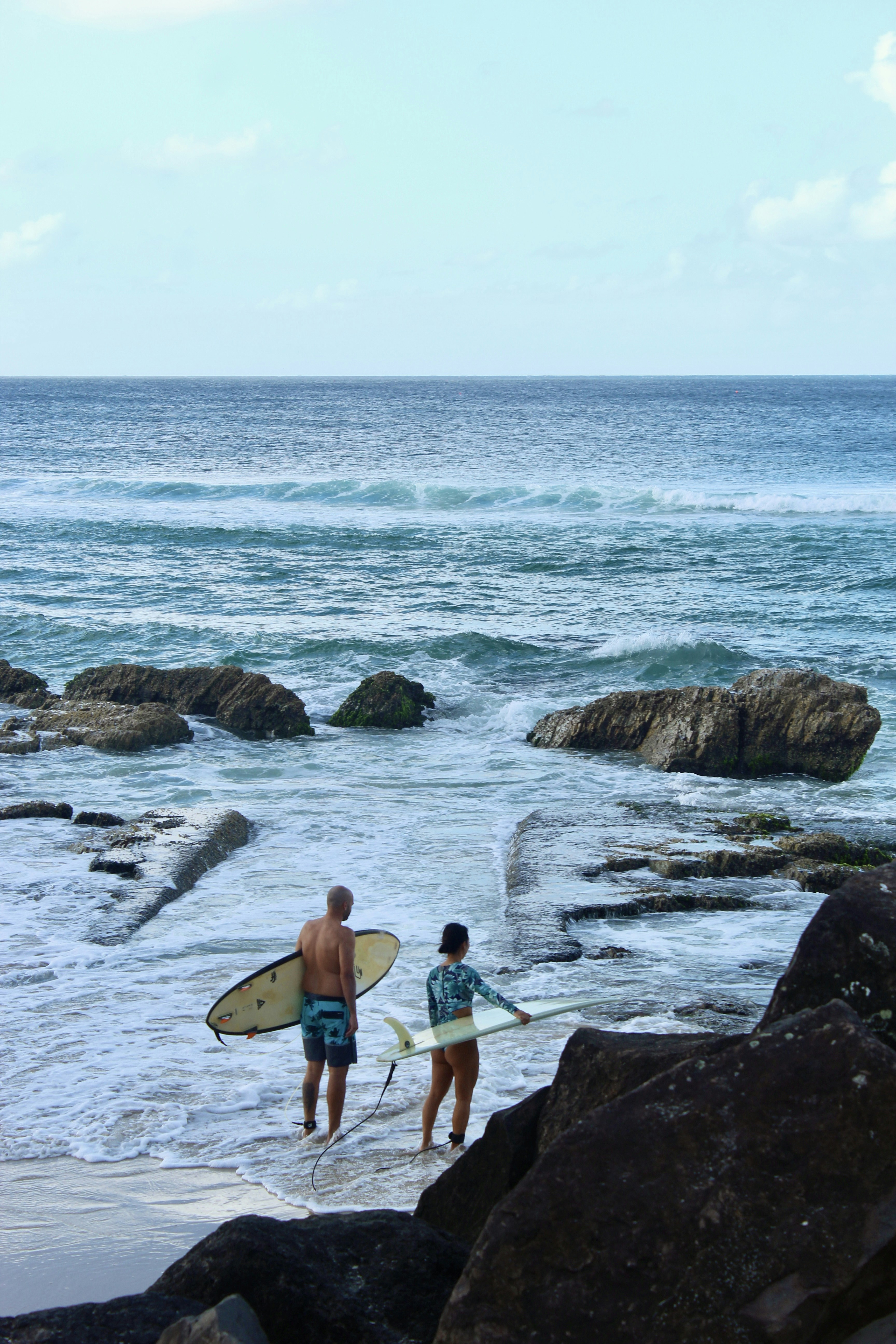 Surfers at the Tidal Barrier: Two Figures Carrying Boards Toward the Sea