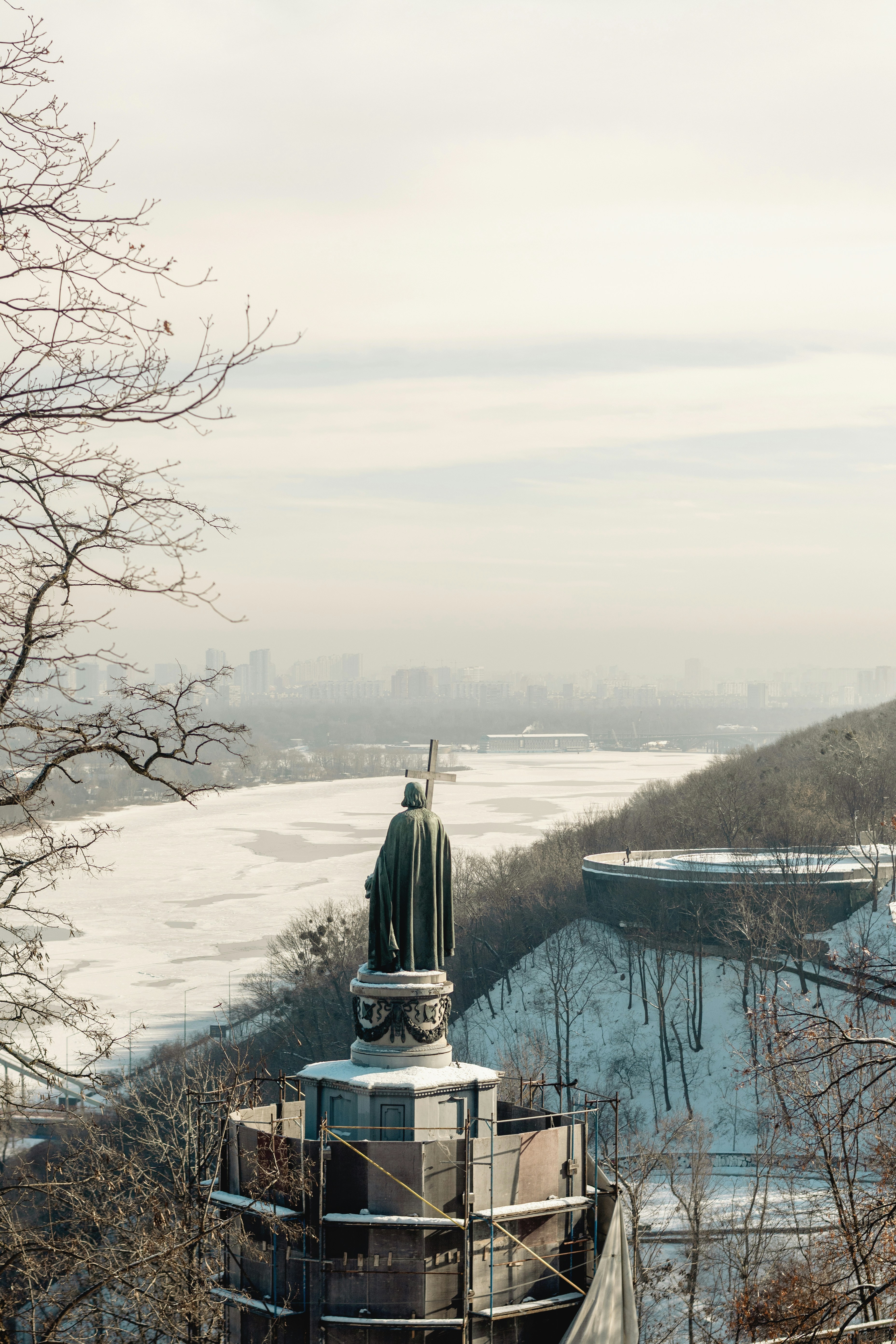 Statue of a cloaked figure overlooking a winter city from a snowy terrace