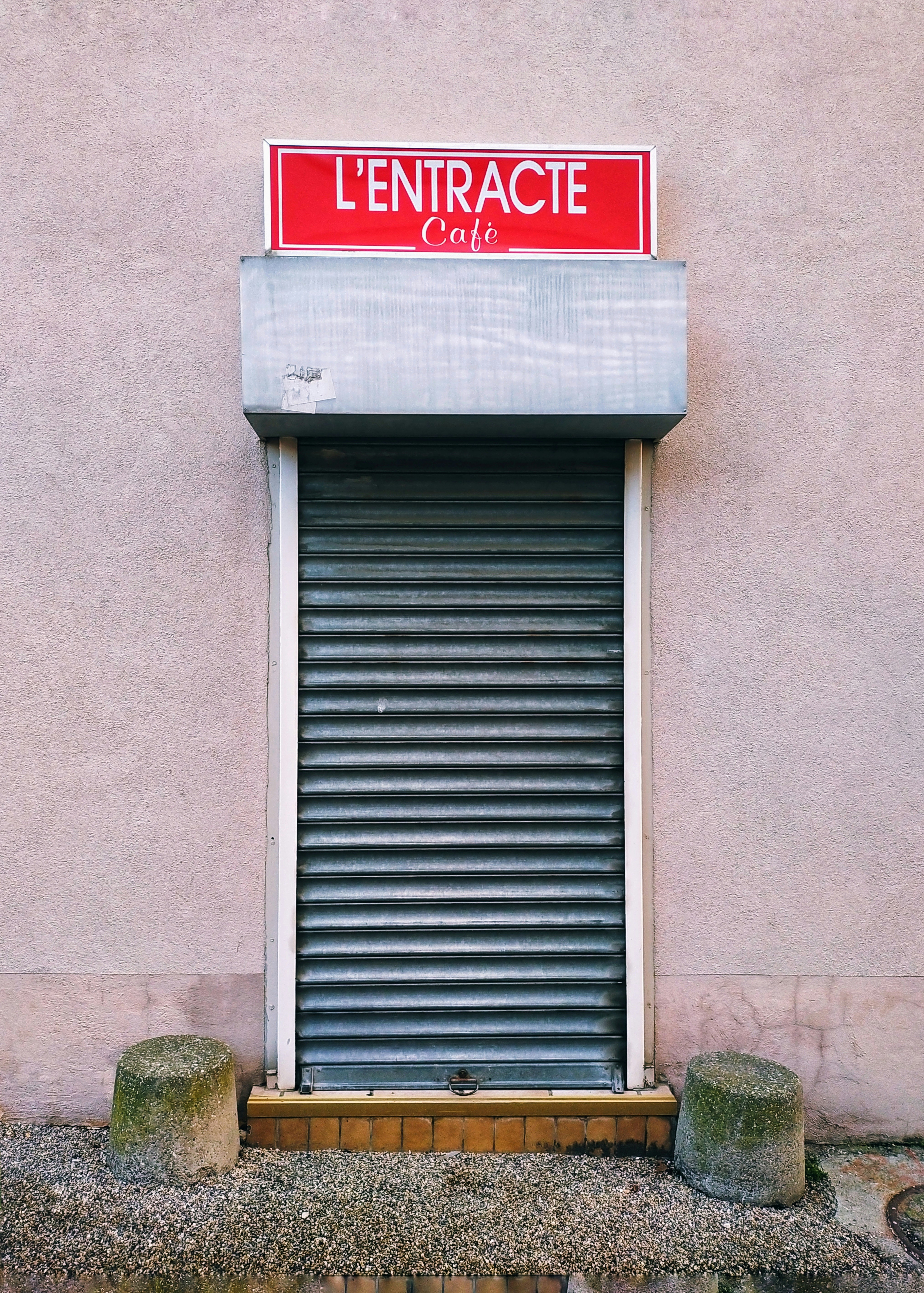 Closed cafe with red l'entracte sign above roller shutter.