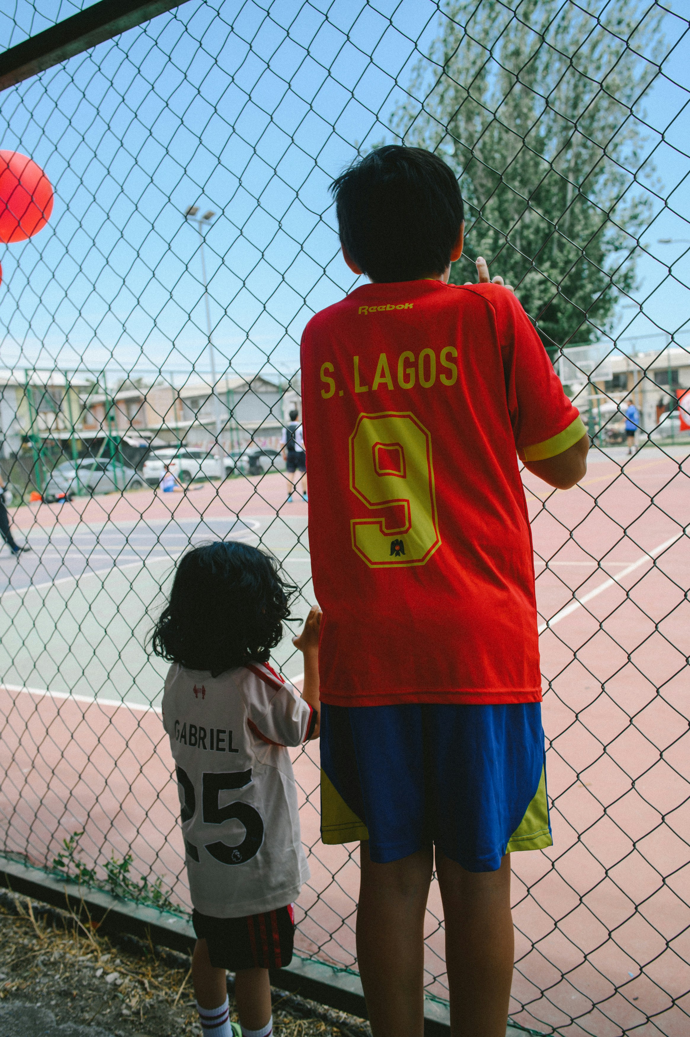 Two Fans at the Chain-Link Stage: A Red Jersey and a Tiny White Jersey Watching the Court