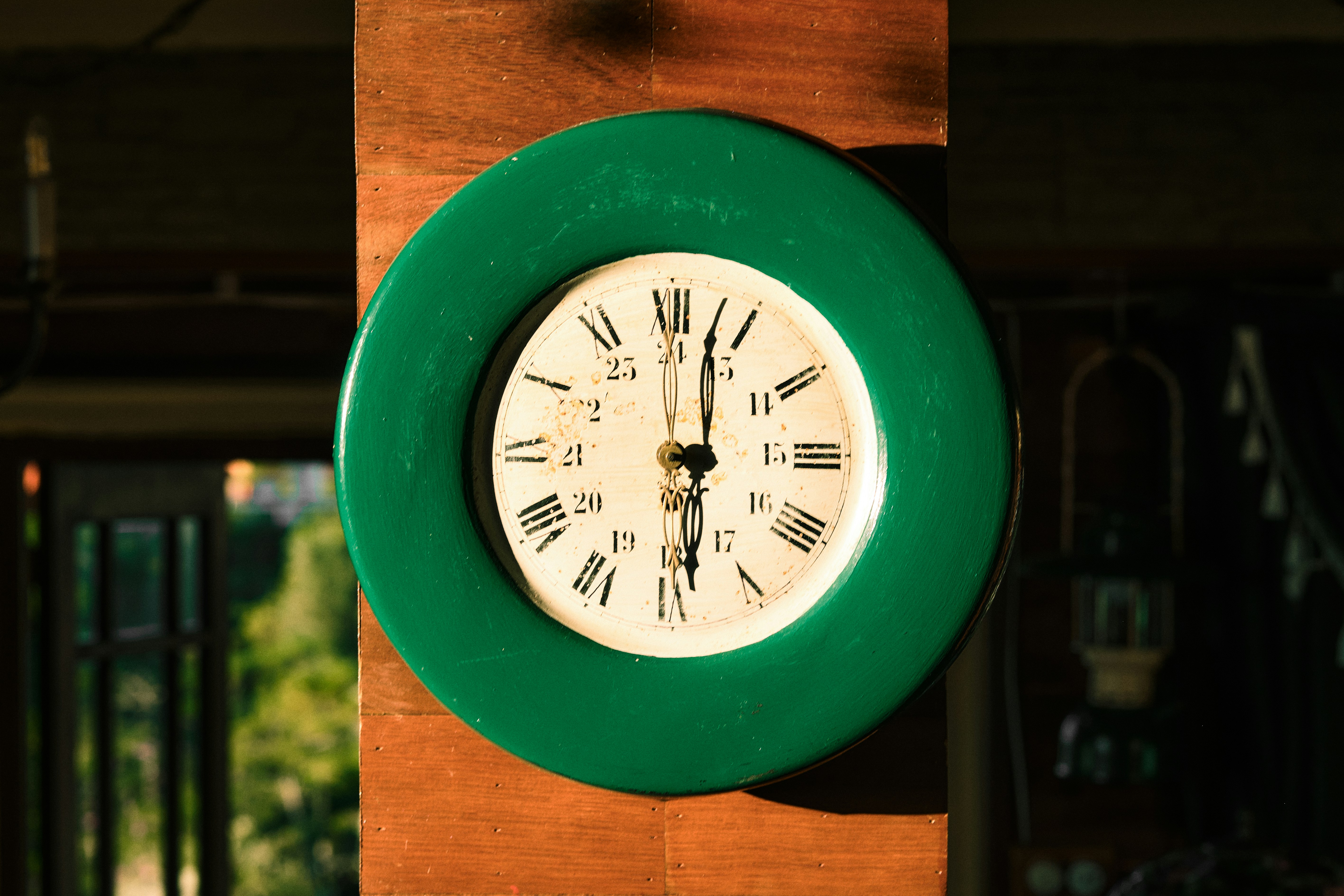 Green-framed clock mounted on a wooden beam, with a blurred interior background, highlighting the timepiece as the focal element.