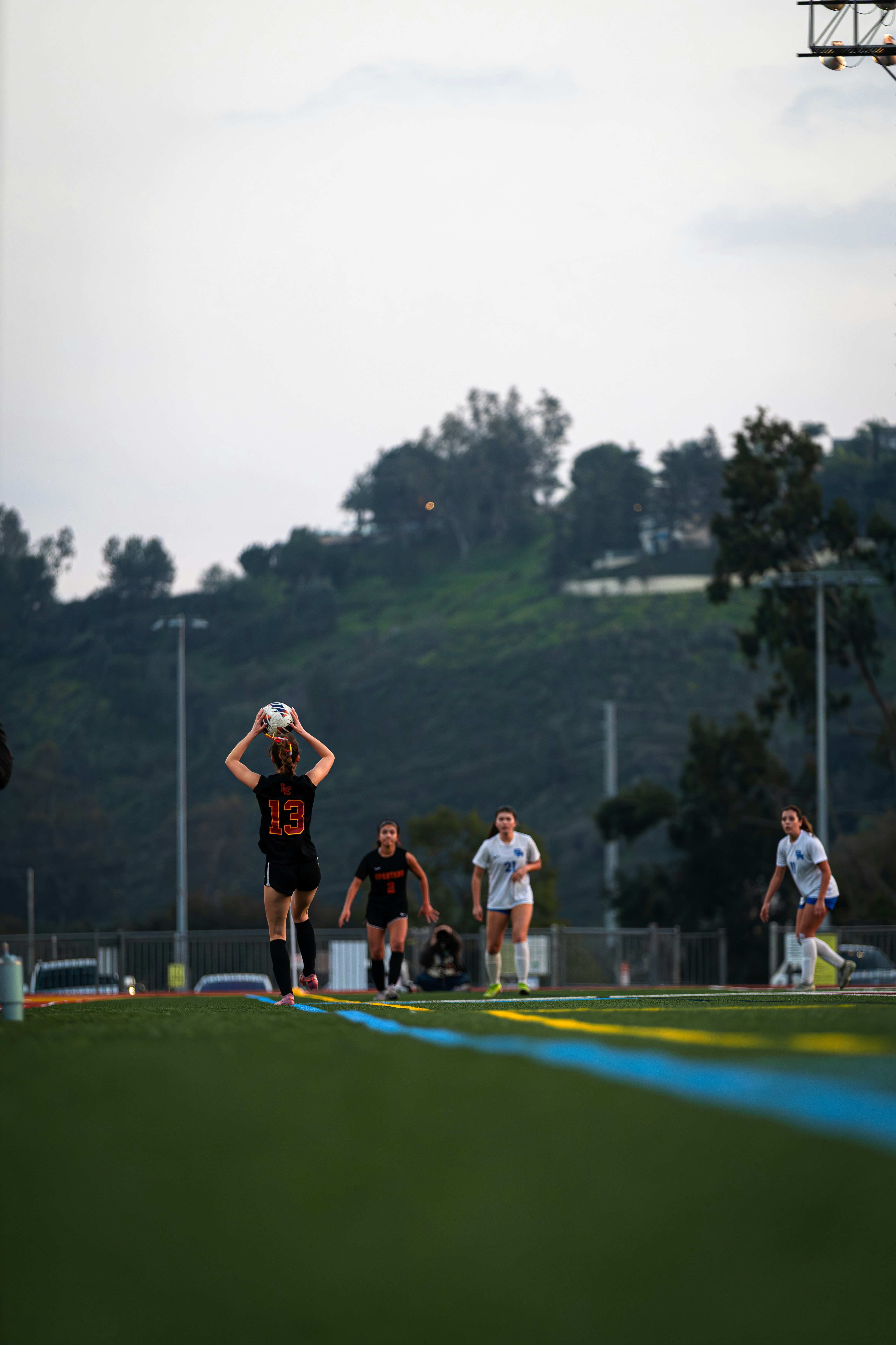 A soccer goalkeeper in black jersey catches a ball near the goal line as teammates and opponents approach on a turf field; a hillside and trees form the backdrop.