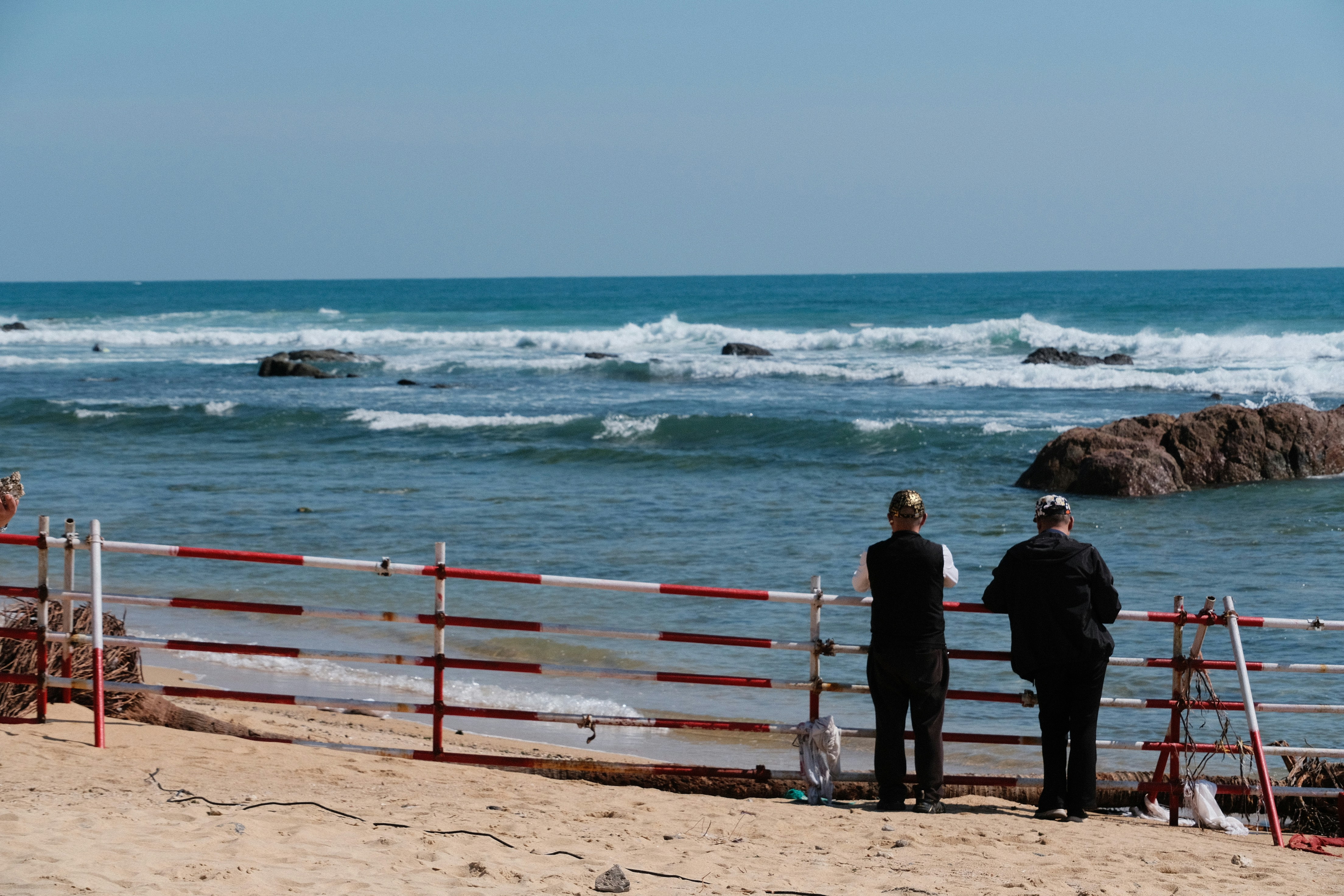 Two shadowed onlookers by a rusting barrier overlook a rolling sea