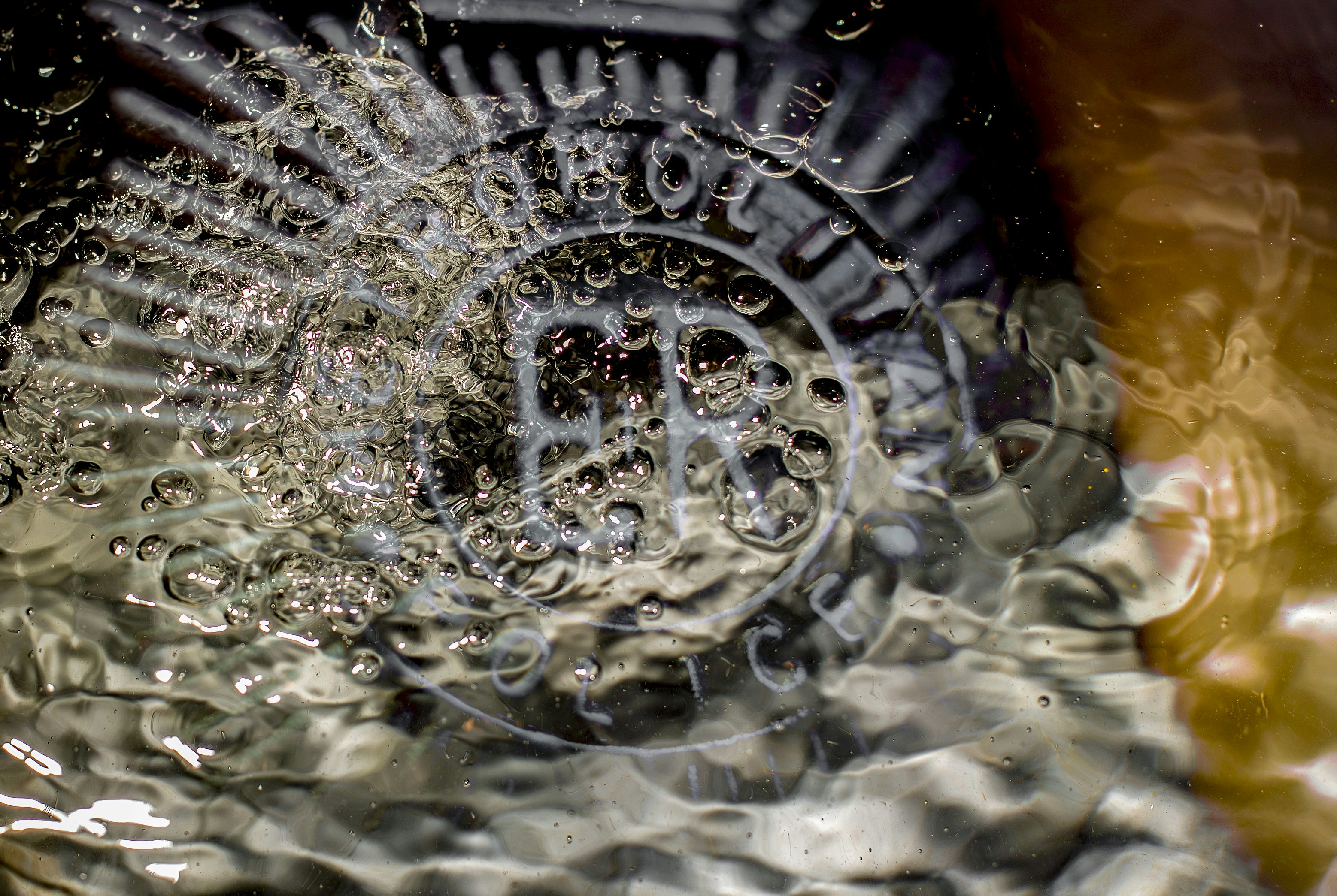 Macro close-up of rippling water or liquid with bubbles and concentric rings; the scene has a glossy, wavy texture suggesting a liquid surface or a glassy artifact. It may resemble a logo watermark or abstract pattern; the depth and gloss imply a photographic capture rather than a painting.