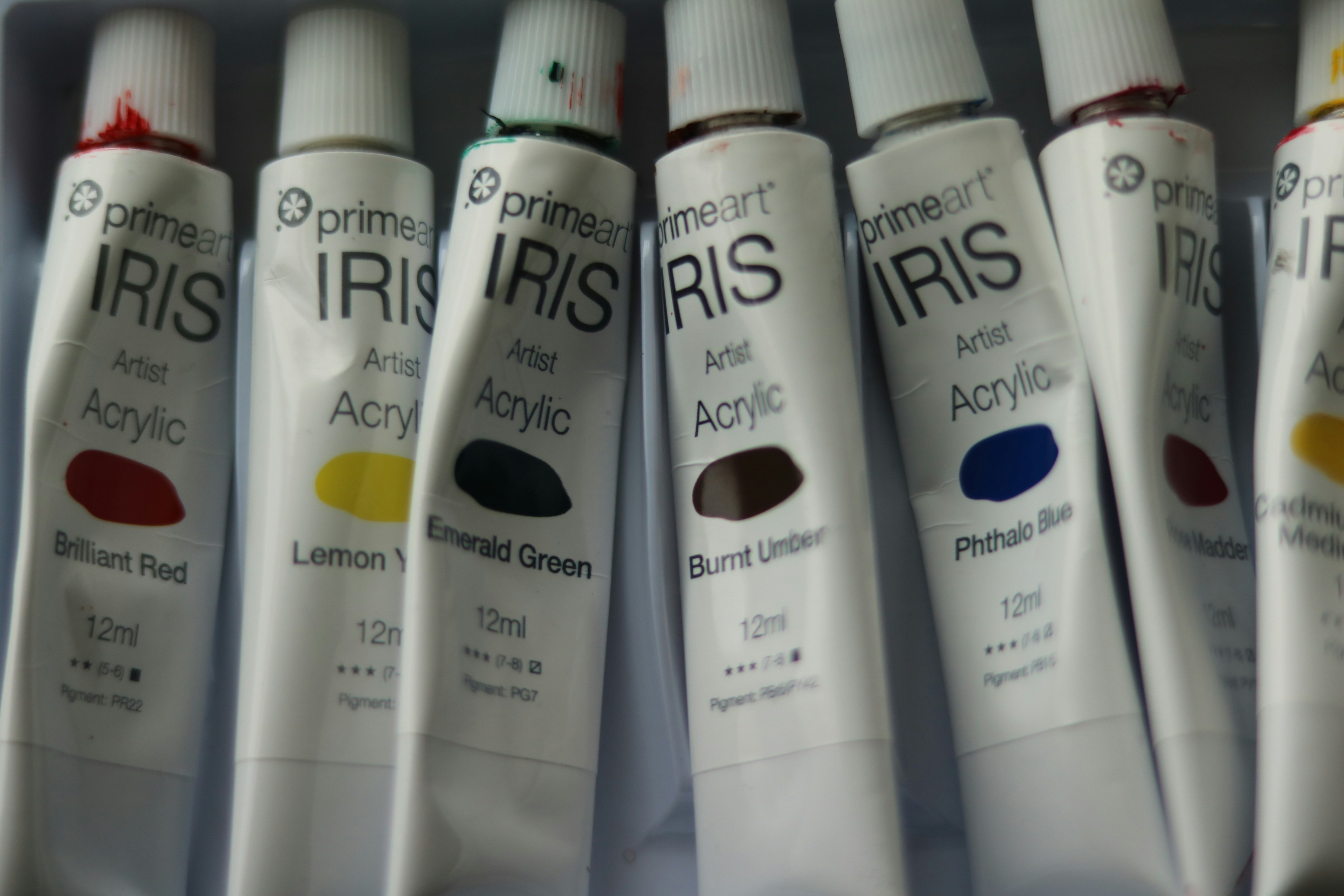 Colorful tubes of Iris acrylic paints arranged in a shallow row, showcasing labels and swatches. The image emphasizes texture and branding in a studio setup.