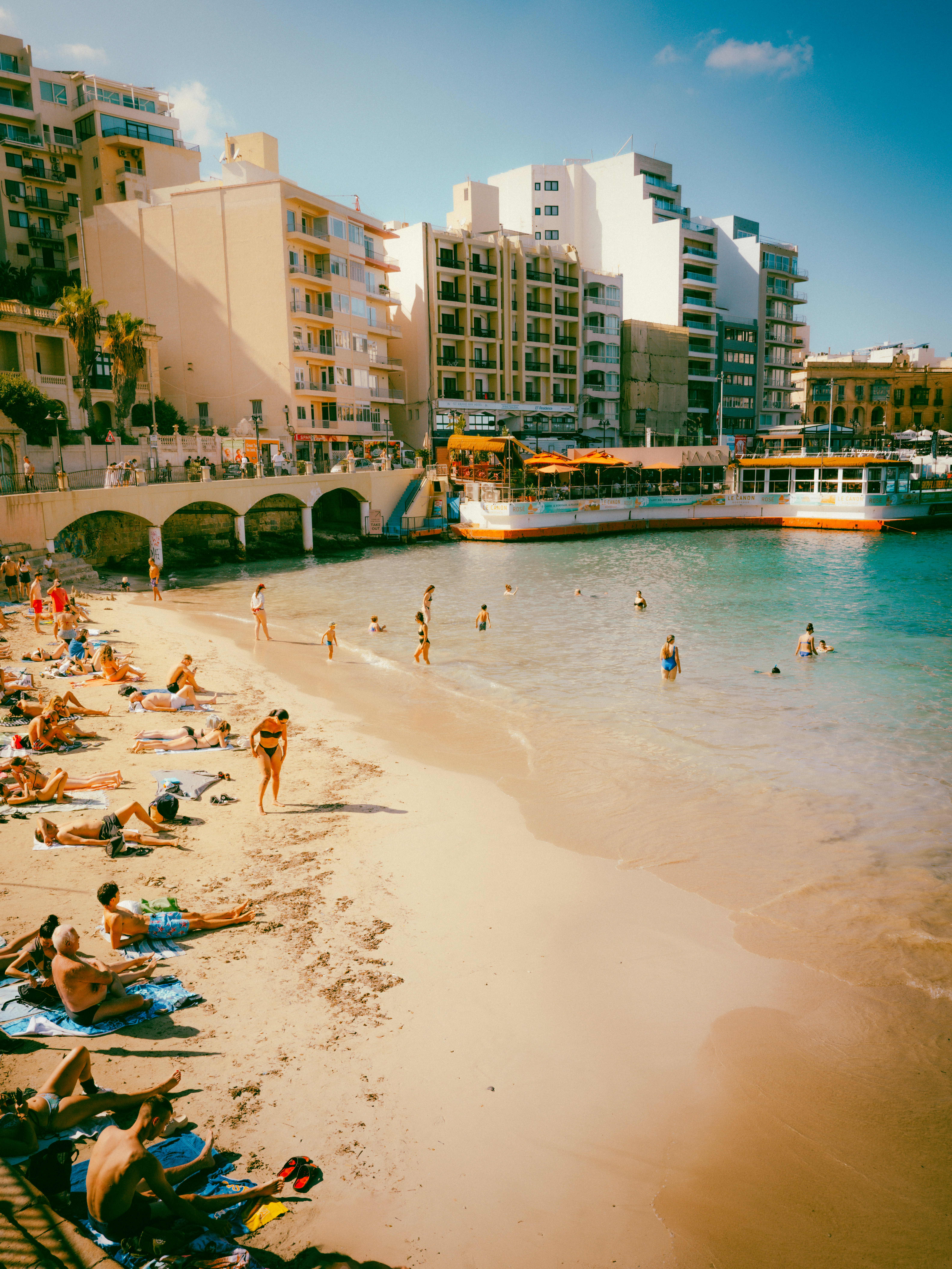 Sunlit Beachfront Mosaic: People, Sand, and Boats by the Waterfront Arcades