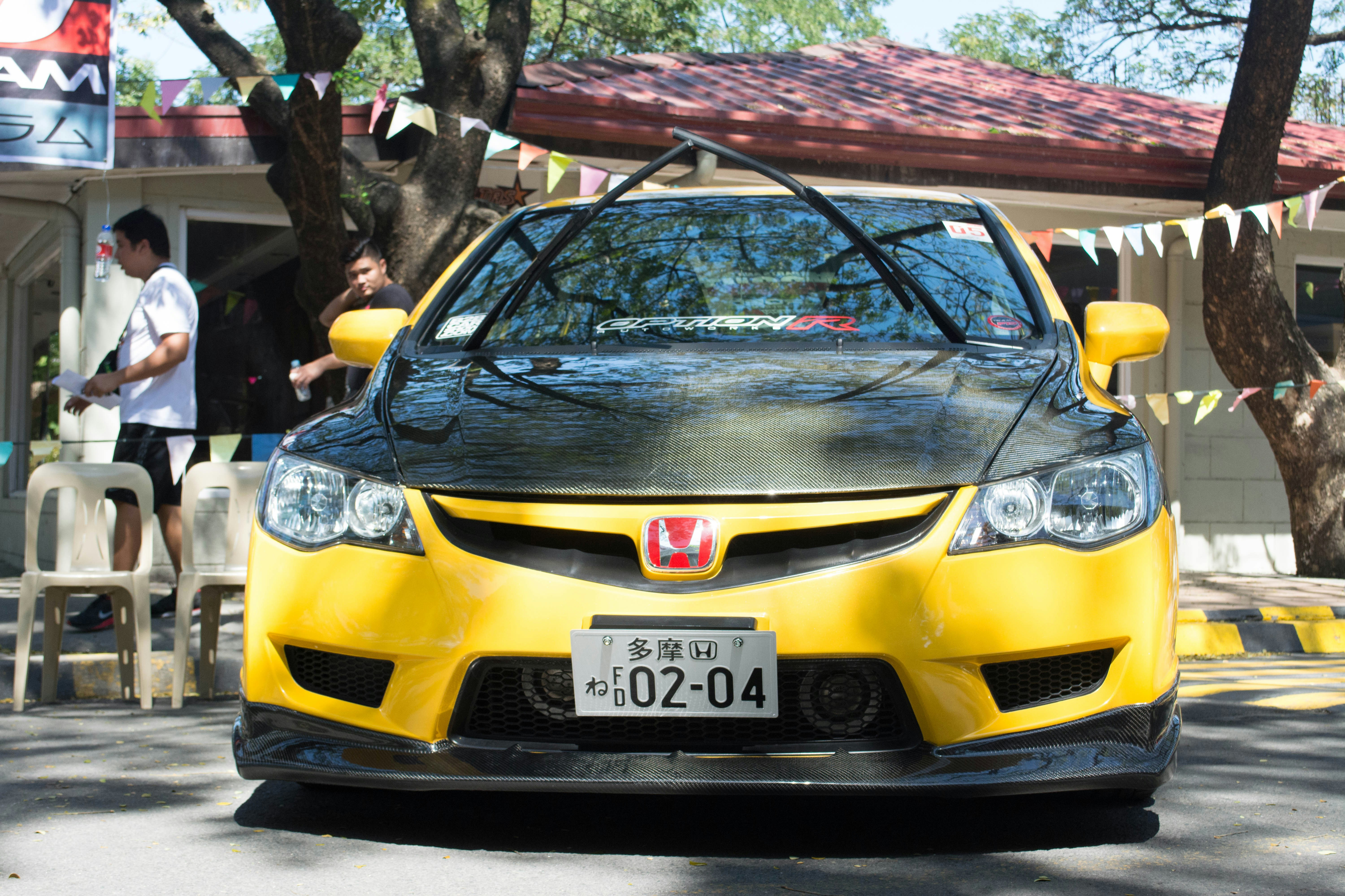 Yellow Honda performance car parked head-on in an outdoor event, license plate visible under a string of pennant banners.