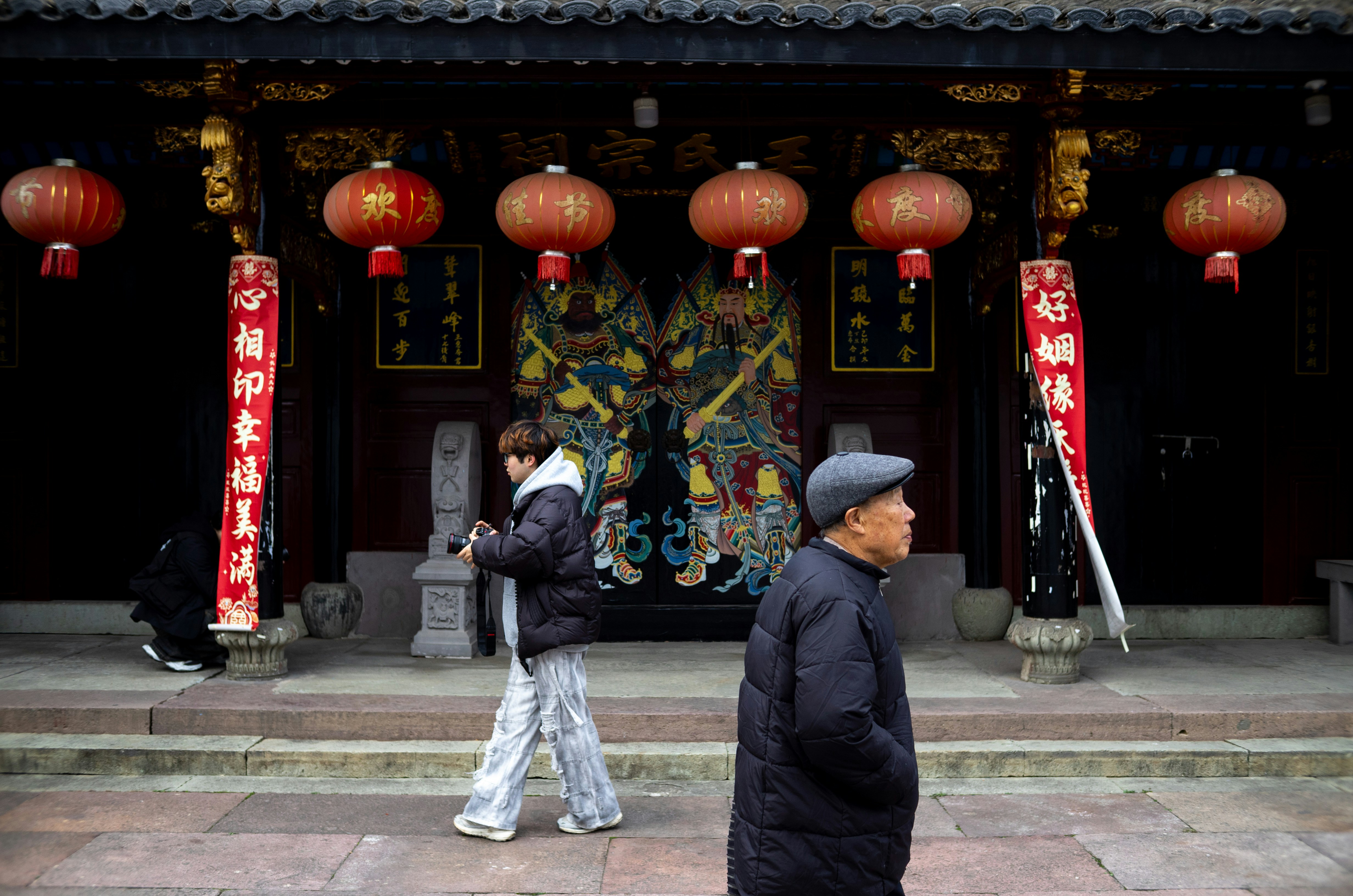 A temple courtyard with red lanterns and a central mural, while two pedestrians pass by in the foreground.