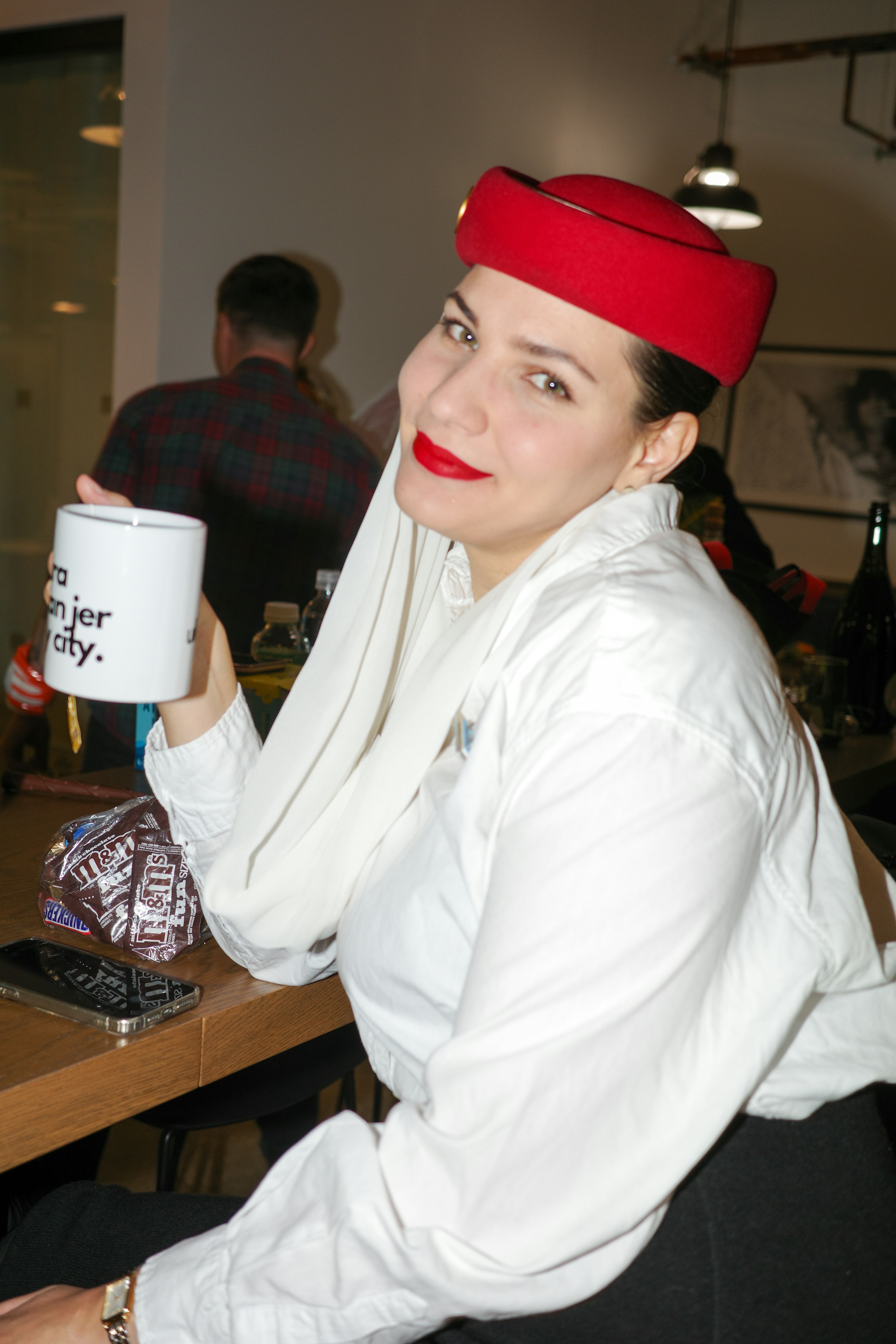 Brightly dressed woman in a red hat and white blouse sits at a cafe counter holding a mug; warm indoor lighting and background diners create a candid social scene.