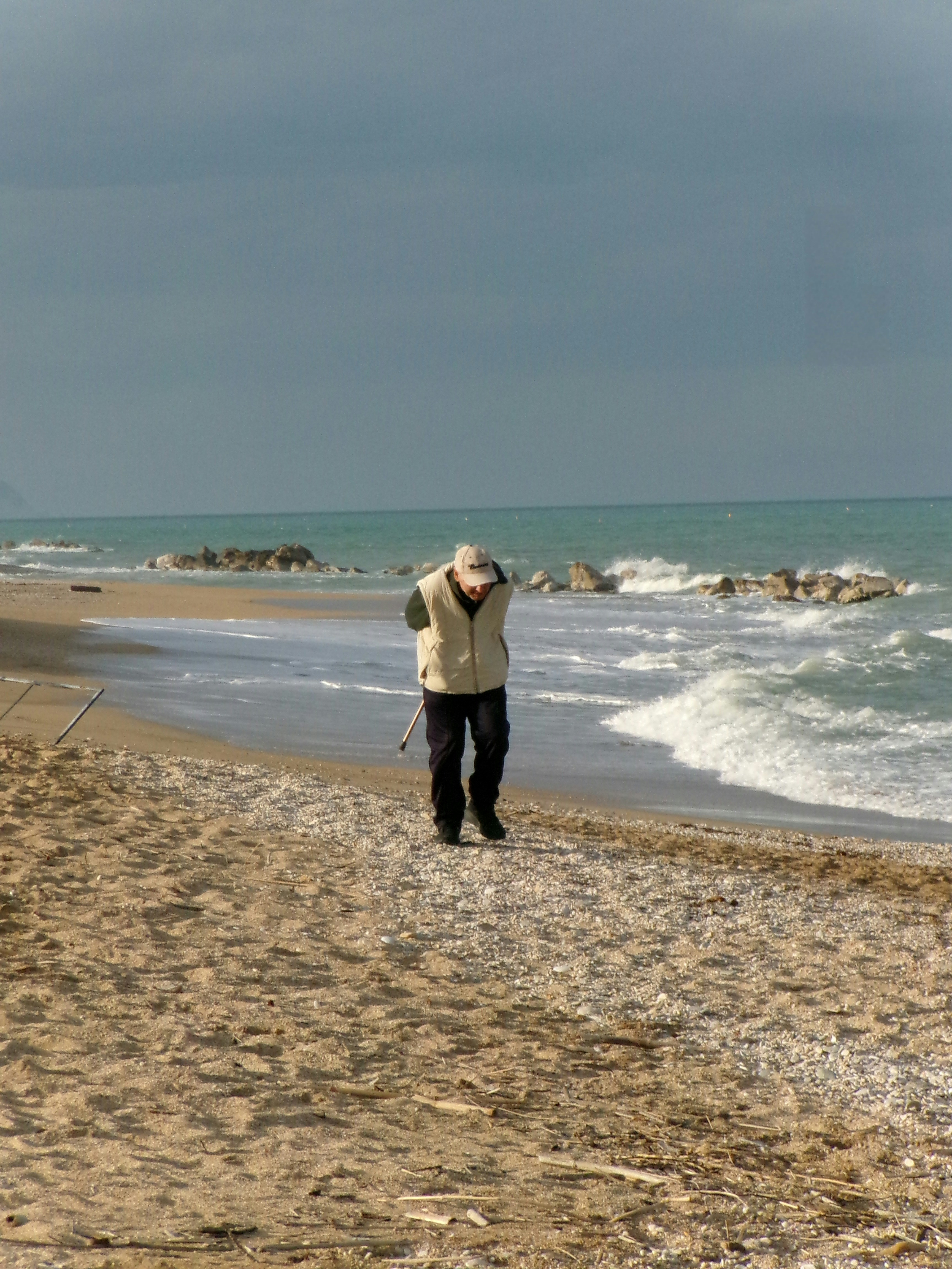 Older man in light vest walking along a pebbly beach with waves crashing nearby; scene captures calm horizon and coastal mood. This is a photographic candid on a shoreline.