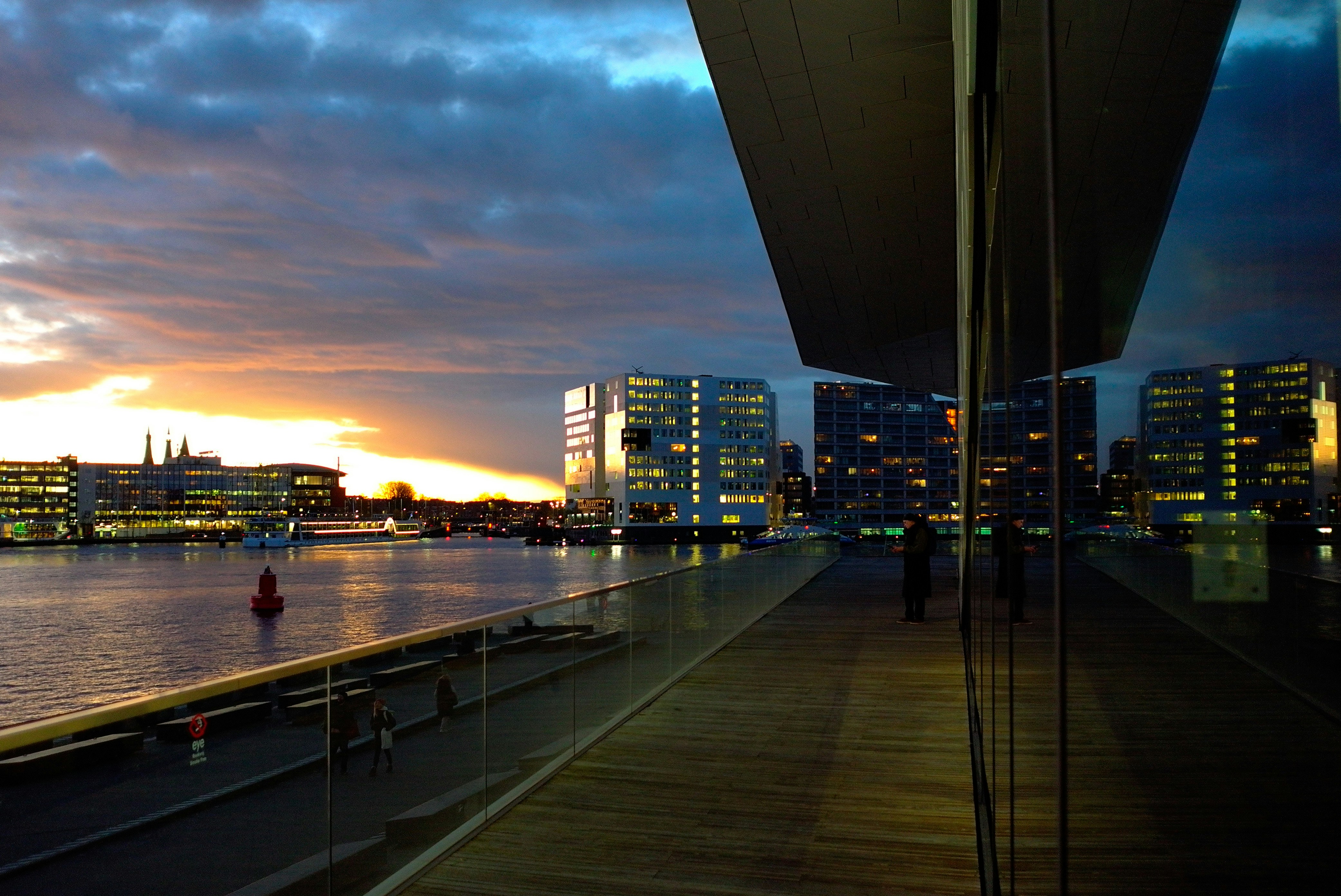 Modern waterfront architecture at sunset with glowing office buildings, river reflections, and a wooden observation deck near the EYE Film Institute