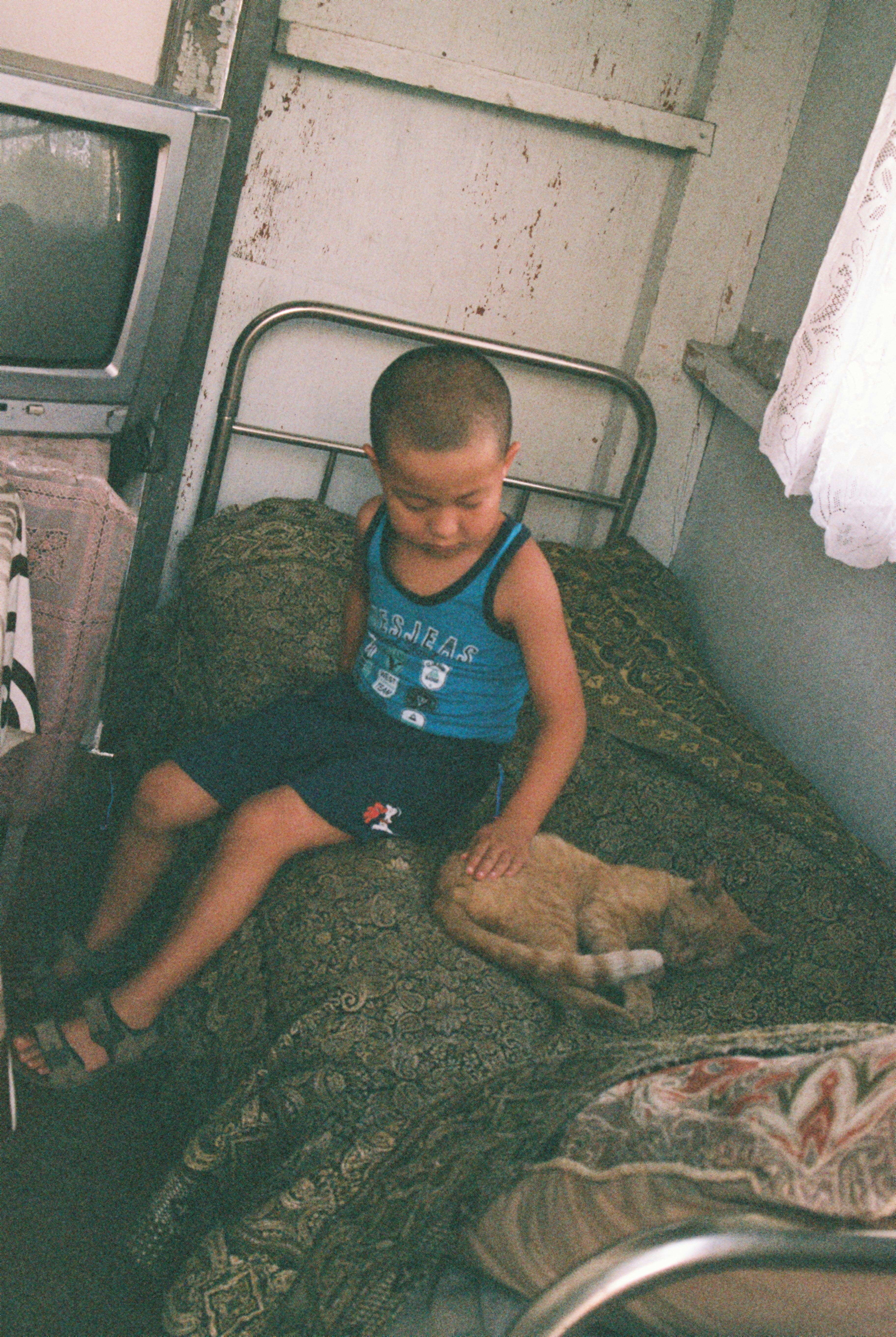 Young boy with shaved head sits on a bed beside an orange cat in a dim, retro room
