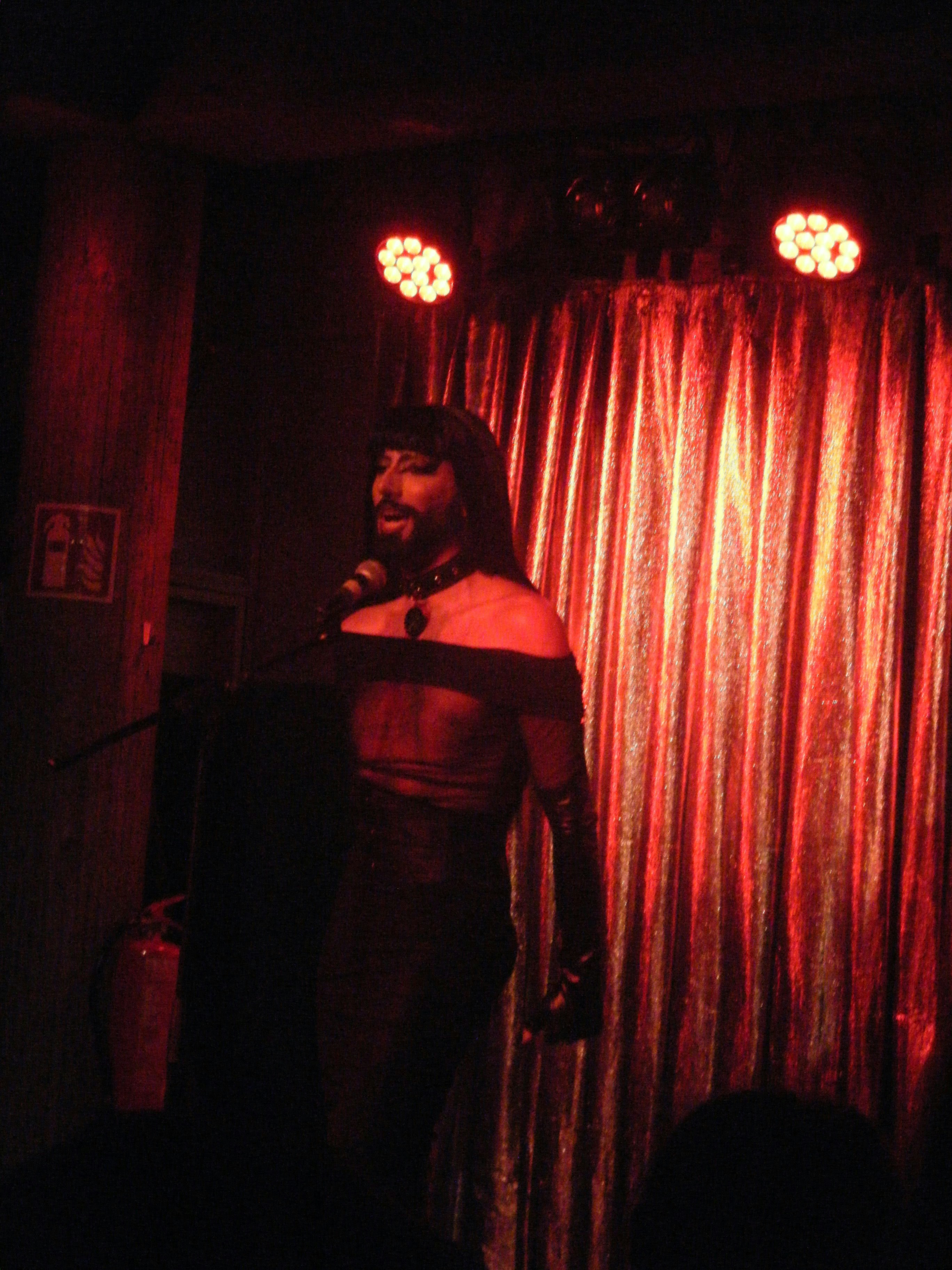 A person in drag performs on stage with red lighting.