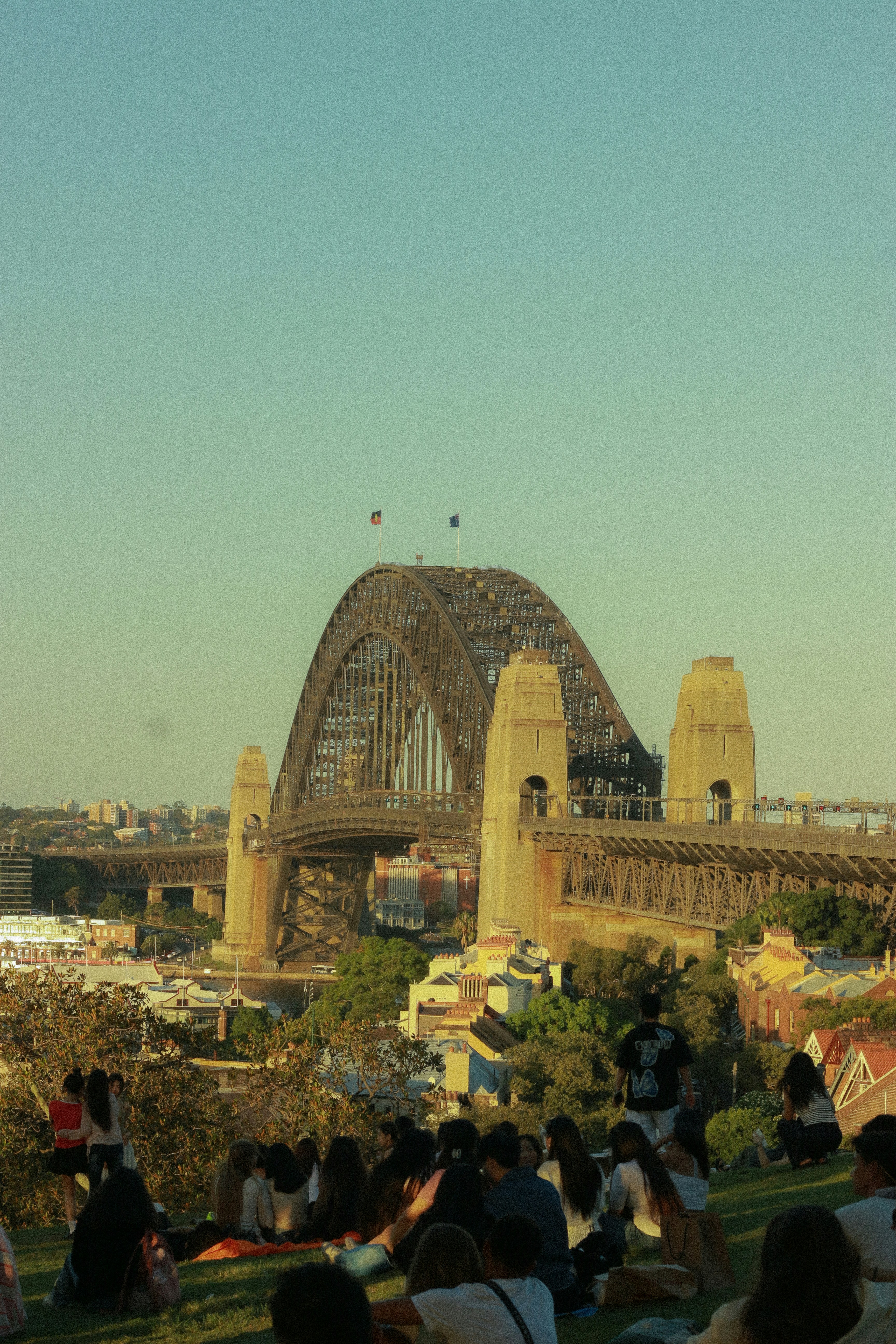 Sunlit Harbour Stone Arc: People Gathered on the Hill Overlooking a Steel-arched Bridge