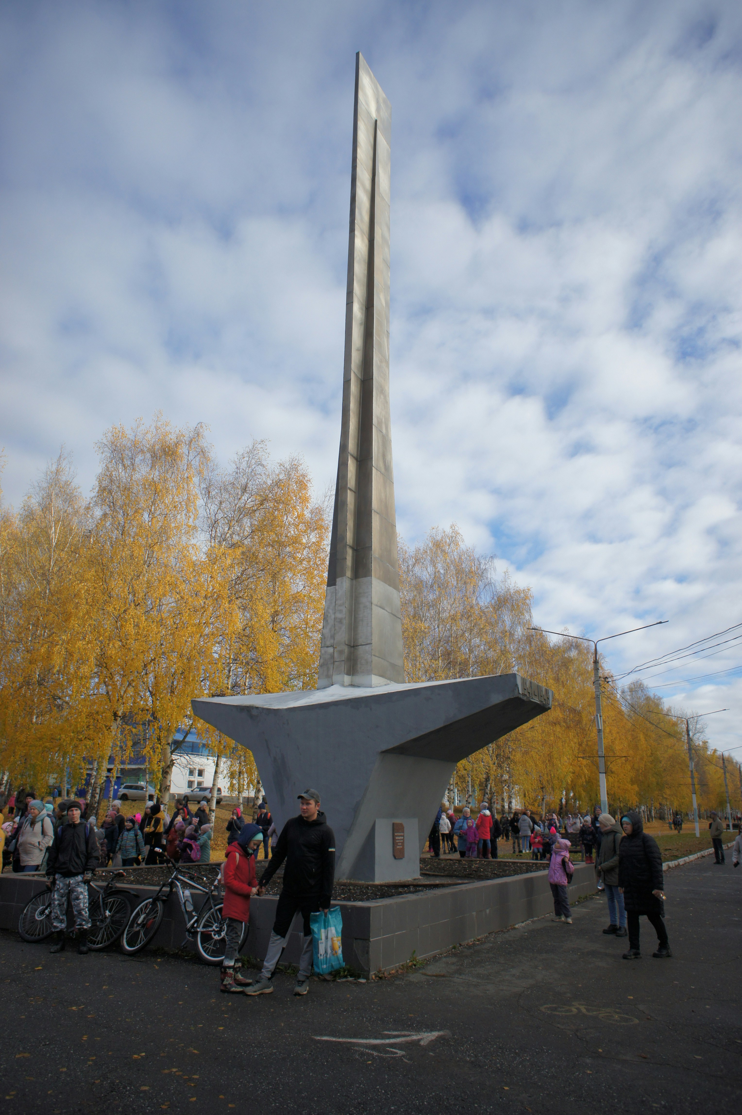 Tall obelisk monument surrounded by autumn trees and people gathered in a plaza with bicycles parked nearby.