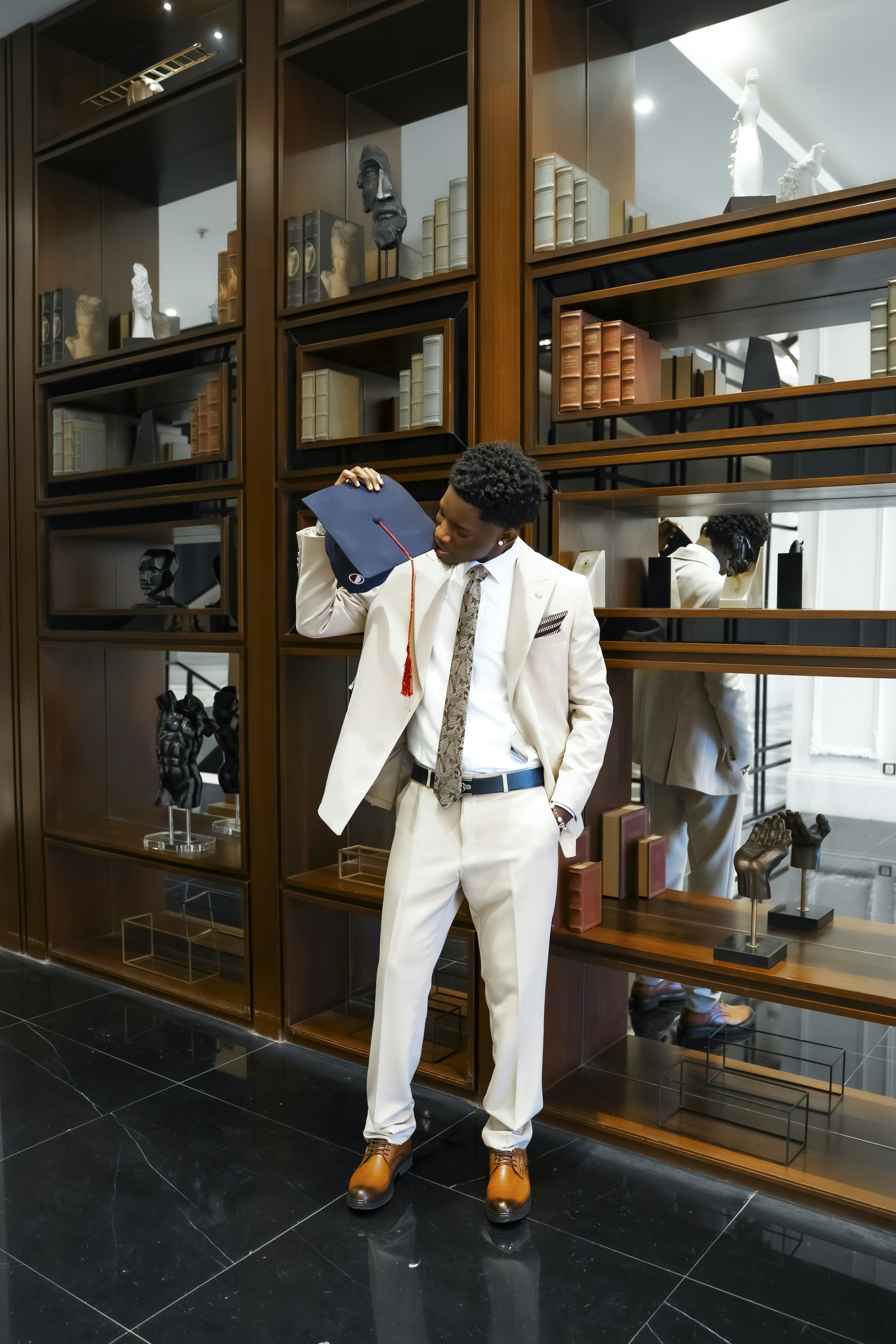 Young man in a white suit holds a graduation cap among wooden shelves filled with books and sculptures; reflective glass panels show his mirrored stance and the refined setting.