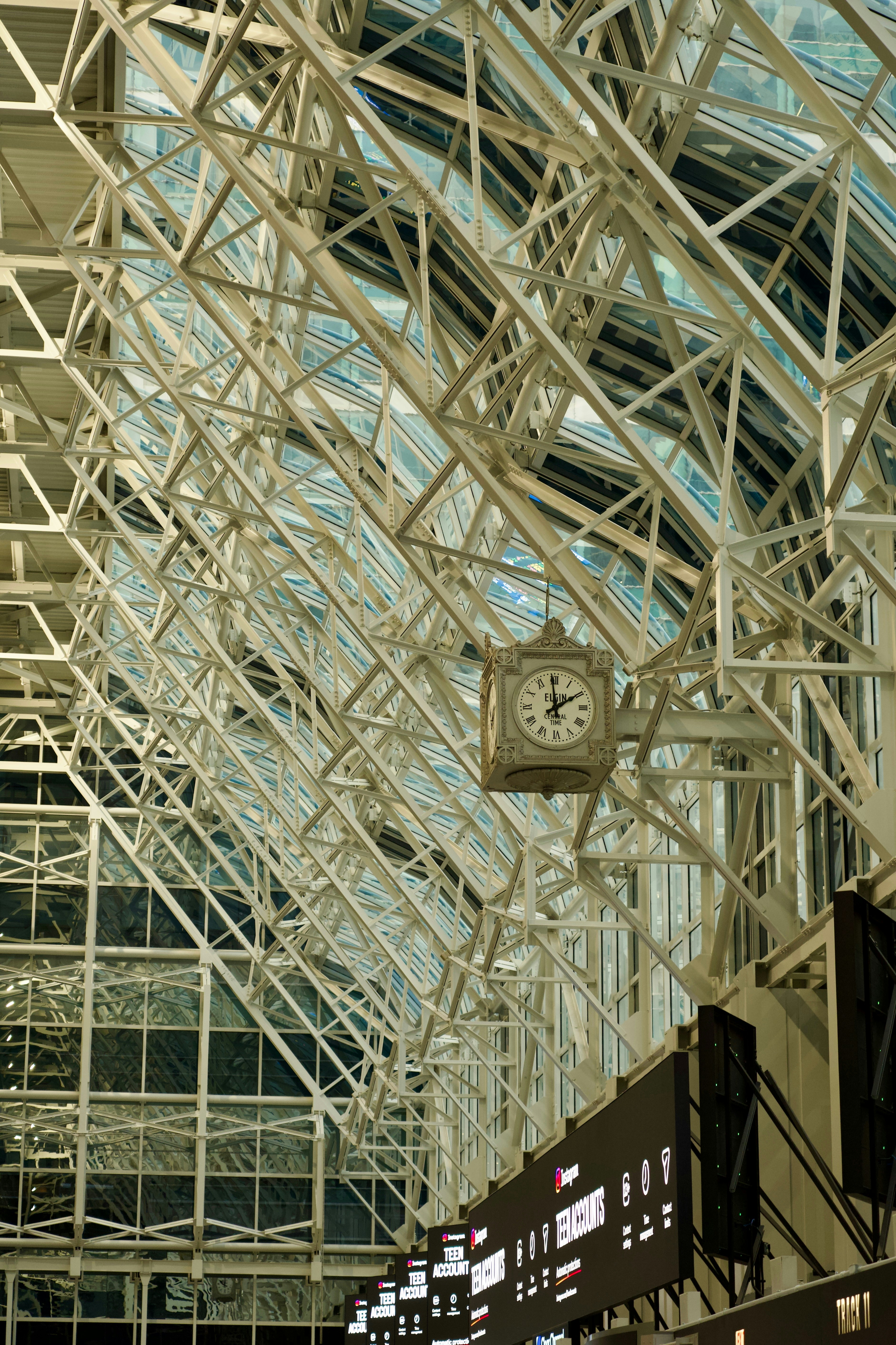 Structural Symphony: A Clock Dangles Amid Crisscrossed Steel in a Glass Atrium