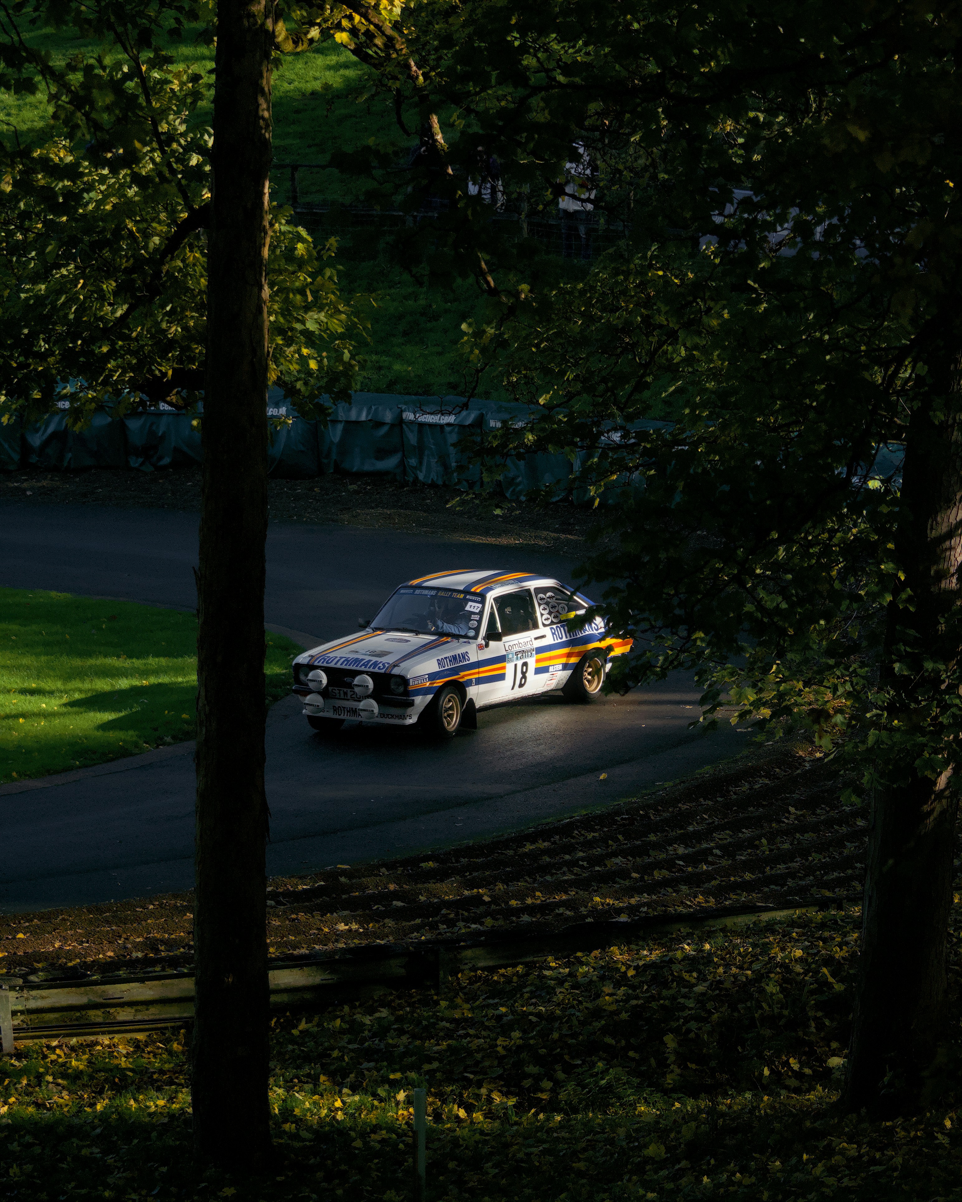 A vintage rally car powers along a sunlit park road, framed by trees and shadows, with its headlights and decals catching the light.