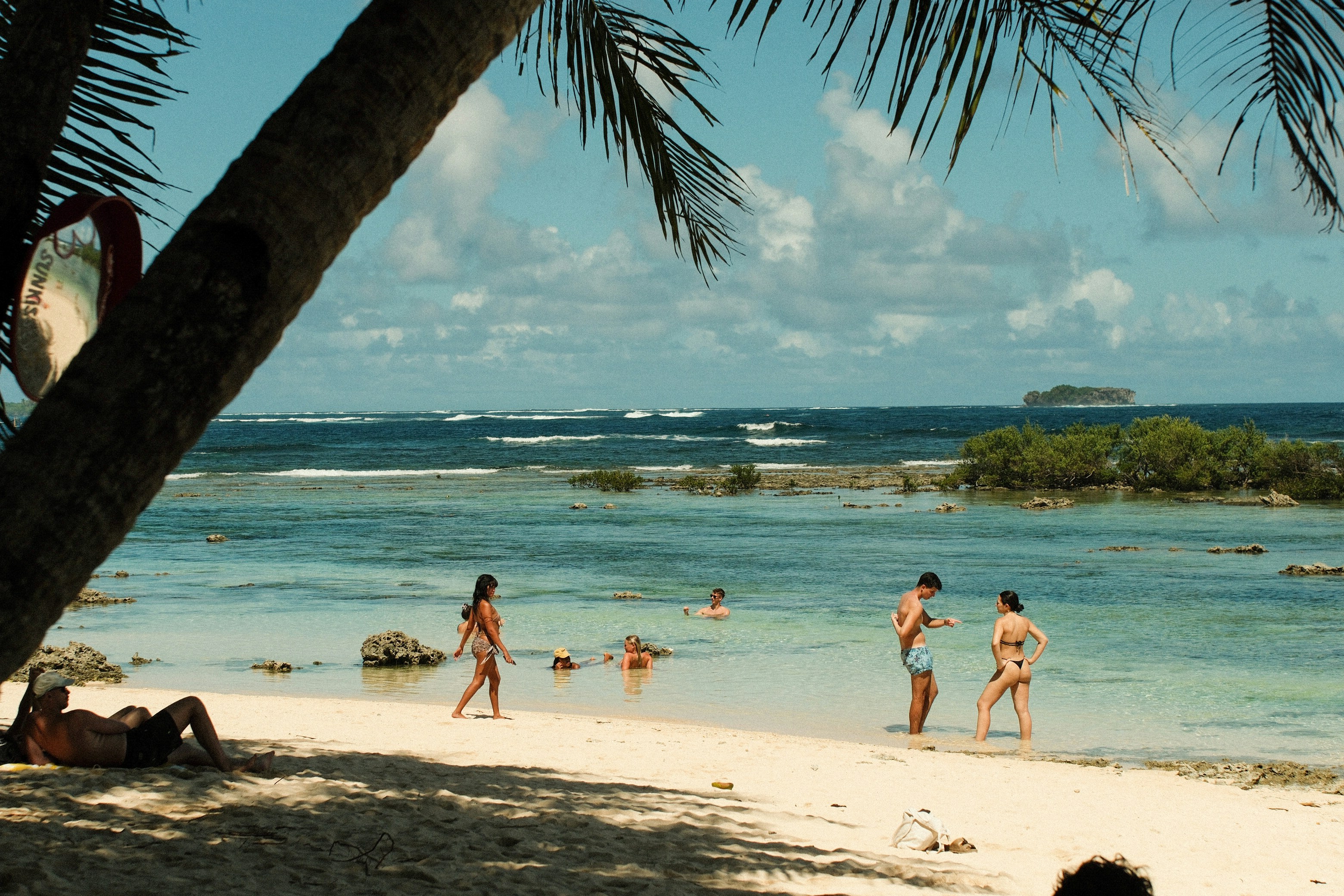Sunlit Beach Scene: Playful Figures in Turquoise Waters under Palm Shade