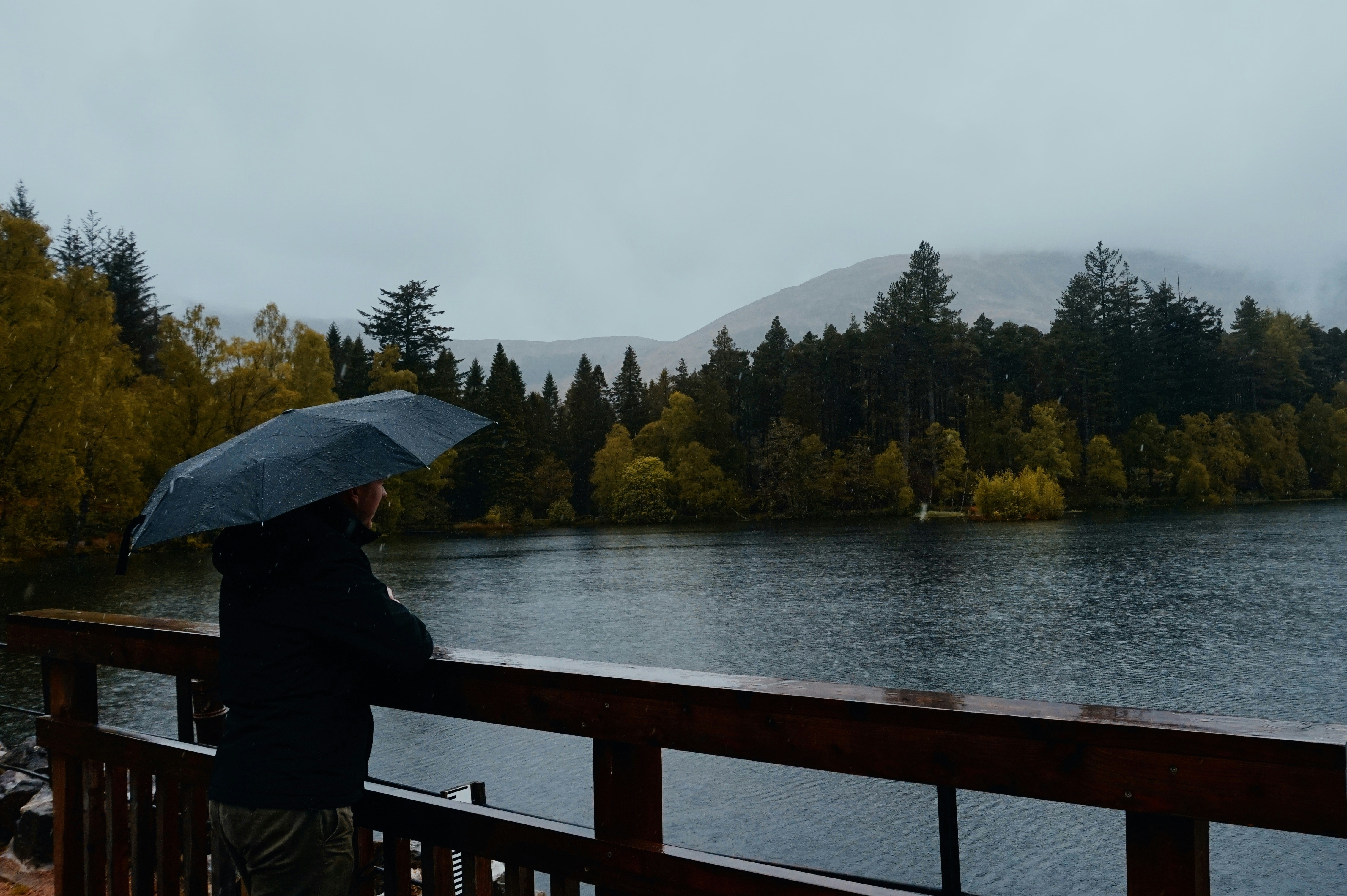 Rainy Riverside: A solitary figure under a blue umbrella by the wooden railing