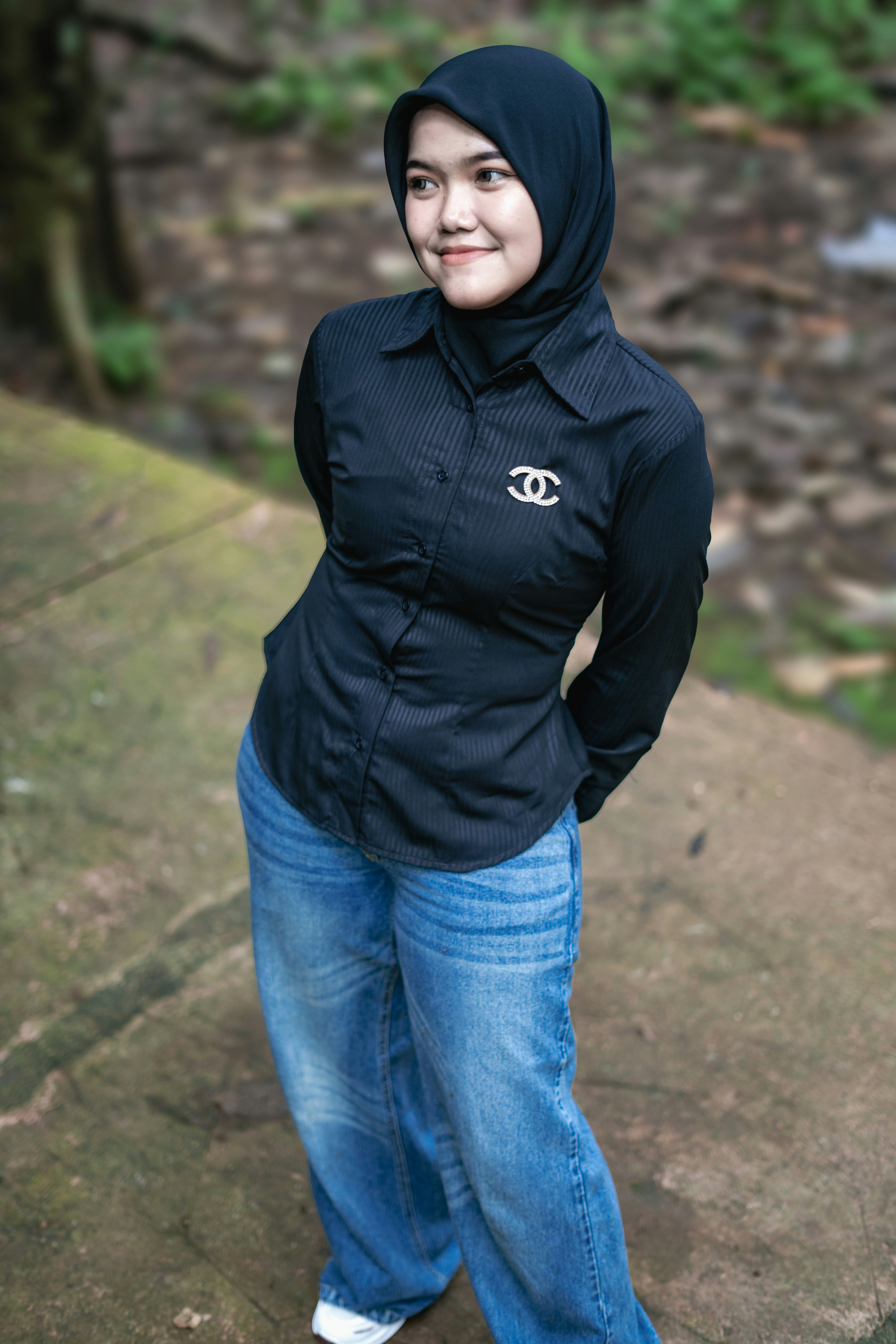 Portrait of a woman in a dark hijab and navy shirt standing outdoors, with a blurred natural background and a Chanel logo on her shirt. The image focuses on the subject's face and upper torso.