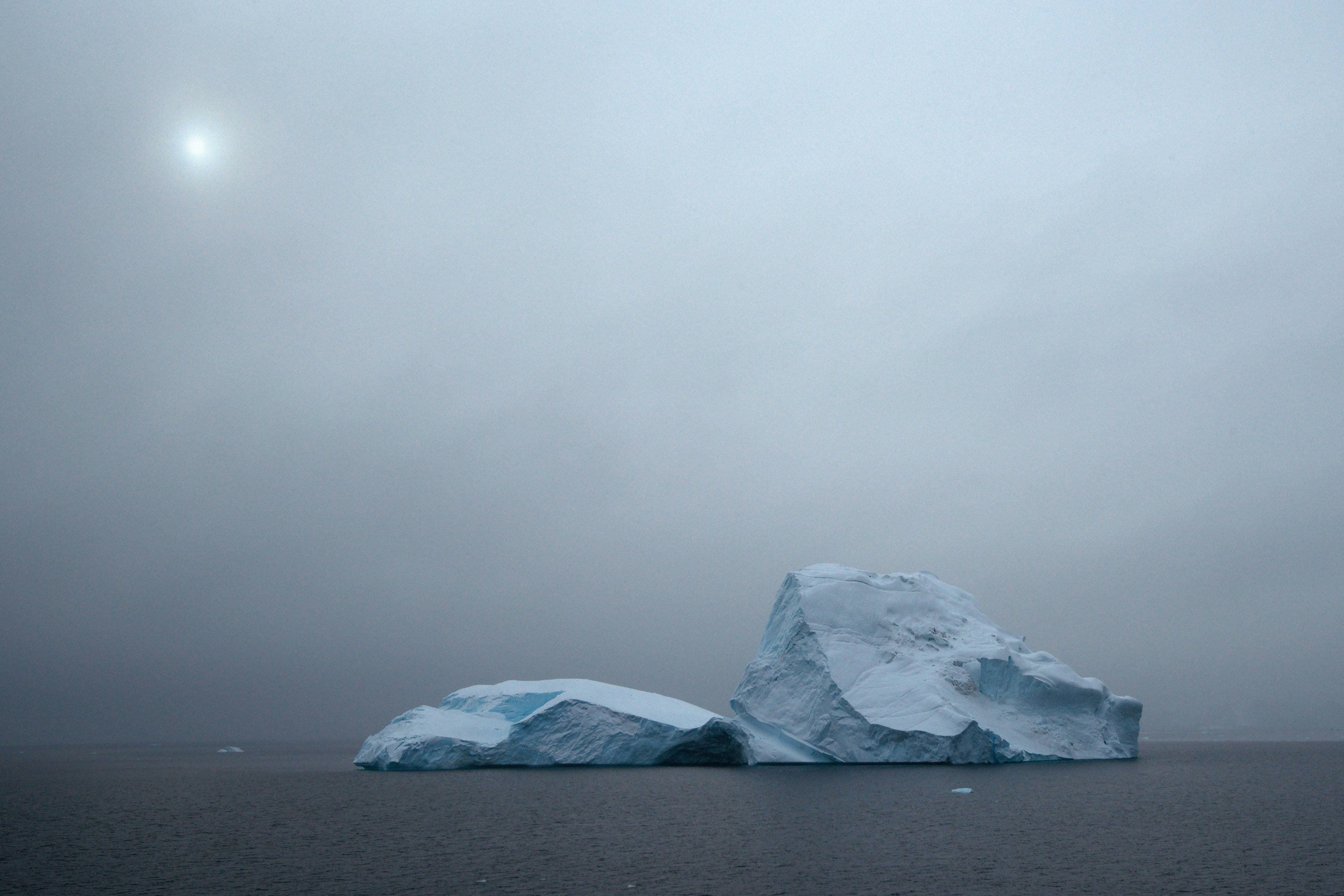 Two icebergs float in a foggy ocean under sun.